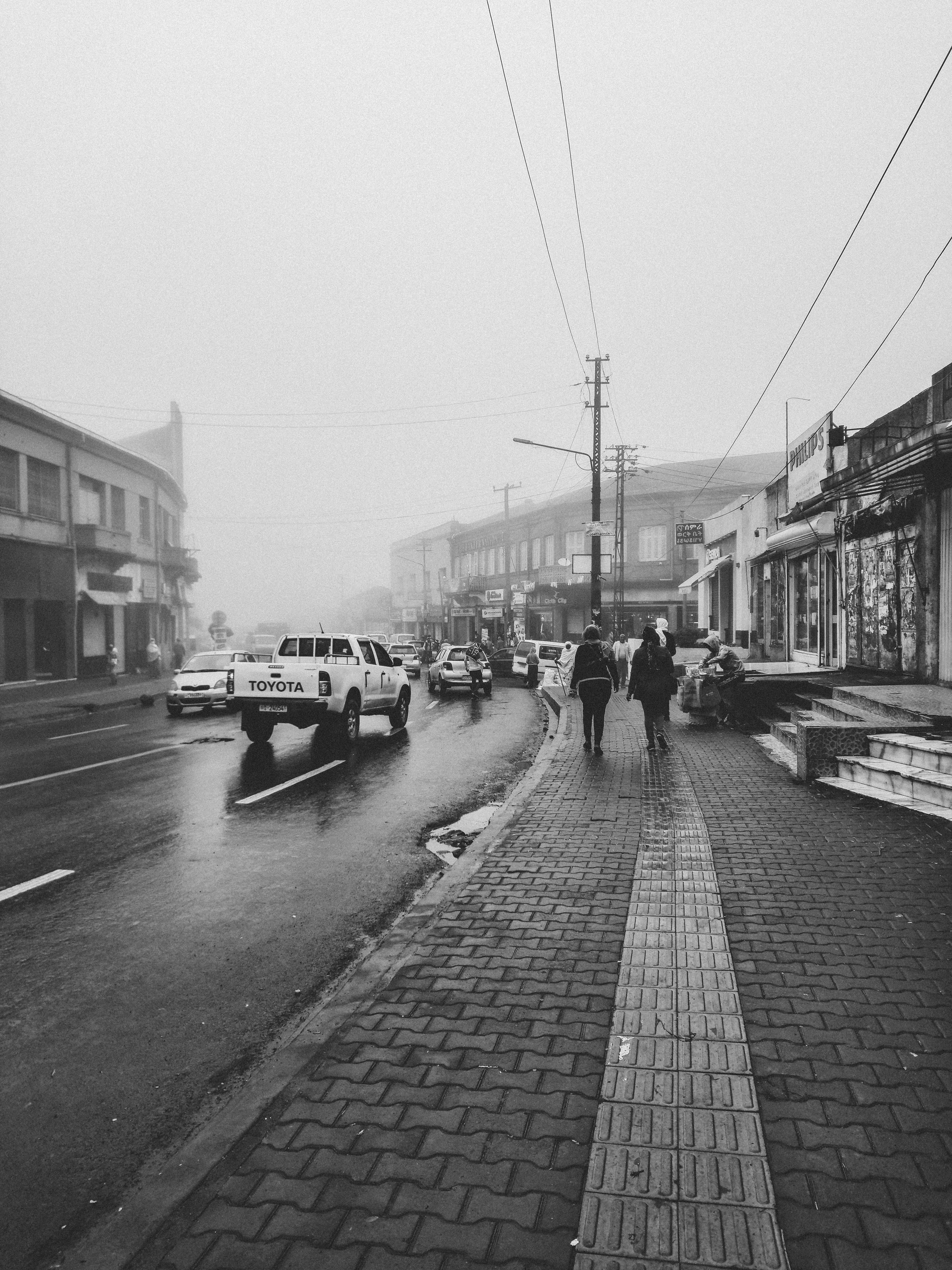 Grayscale street scene with pedestrians along a wet sidewalk beside storefronts while cars pass through a foggy, quiet town.