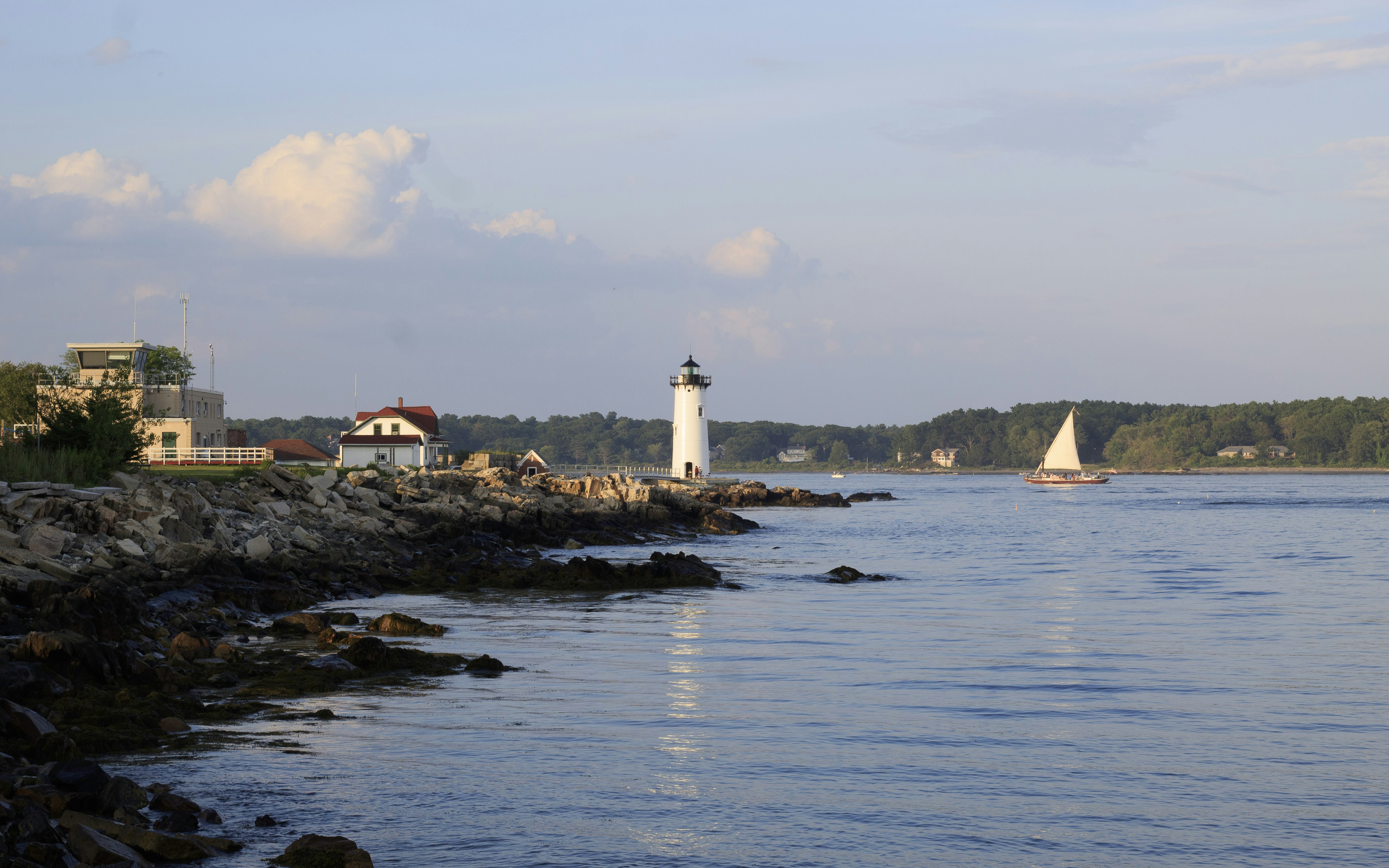 Lighthouse standing on rocky shoreline with a sailboat gliding in the calm sea at sunset.