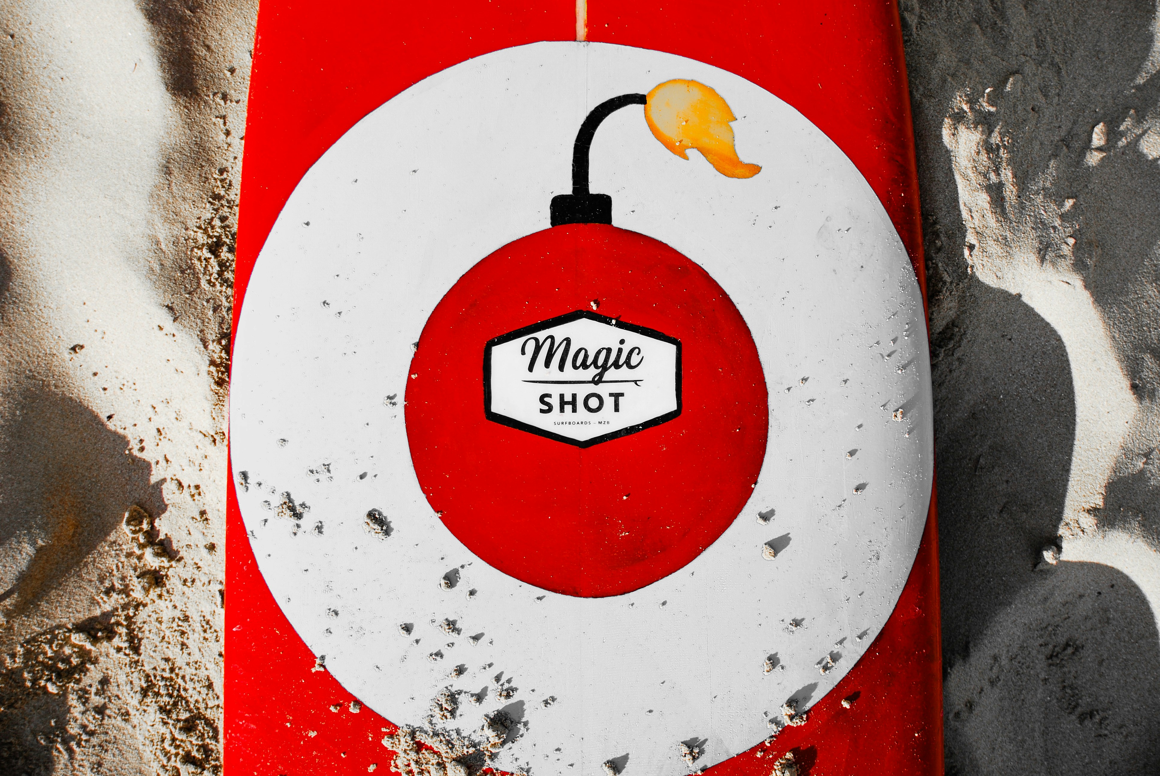 A vibrant surfboard featuring a playful logo with the text 'Magic Shot' surrounded by a red and white design. Sand textures enhance the beach vibe.