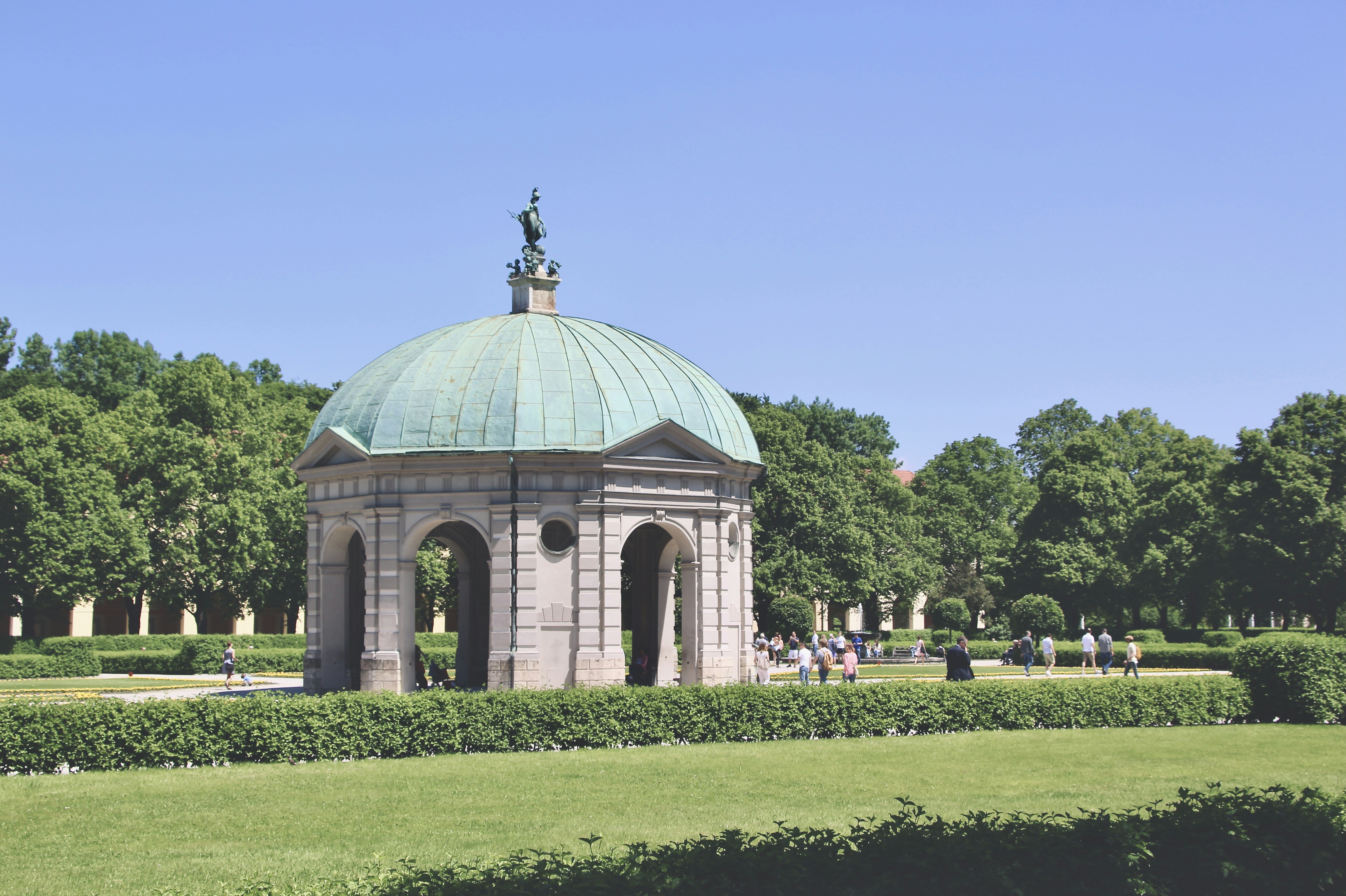 Historical pavilion with a green dome surrounded by lush gardens and visitors enjoying the scenery.