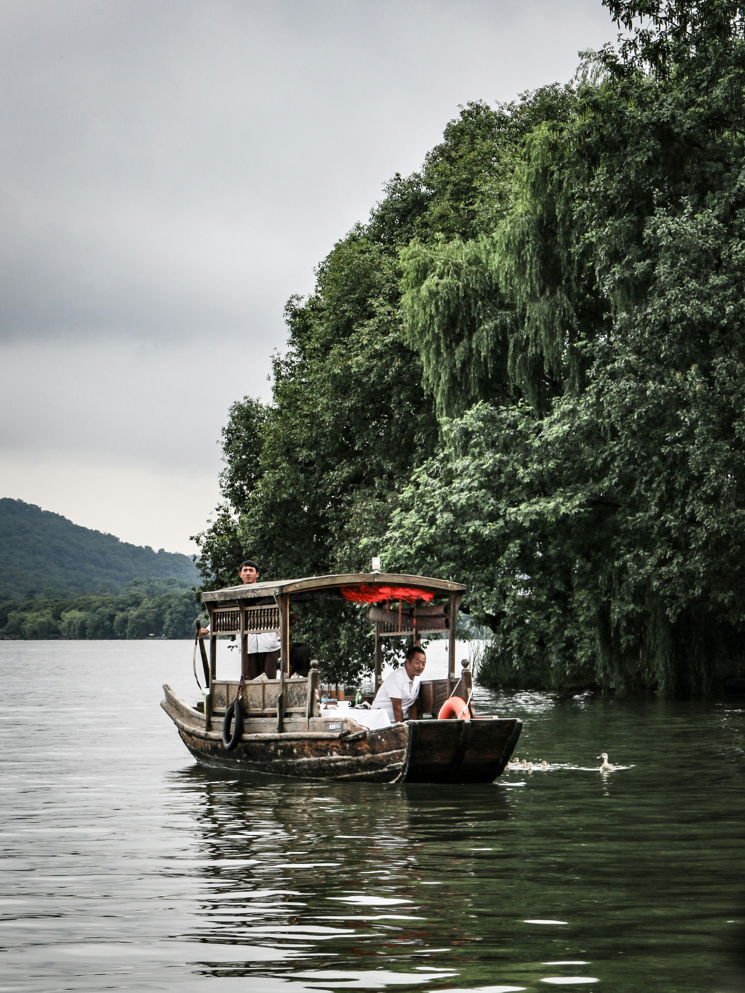 Traditional wooden boat navigating a tranquil lake surrounded by lush greenery and distant hills. Two fishermen engage in their daily routine.