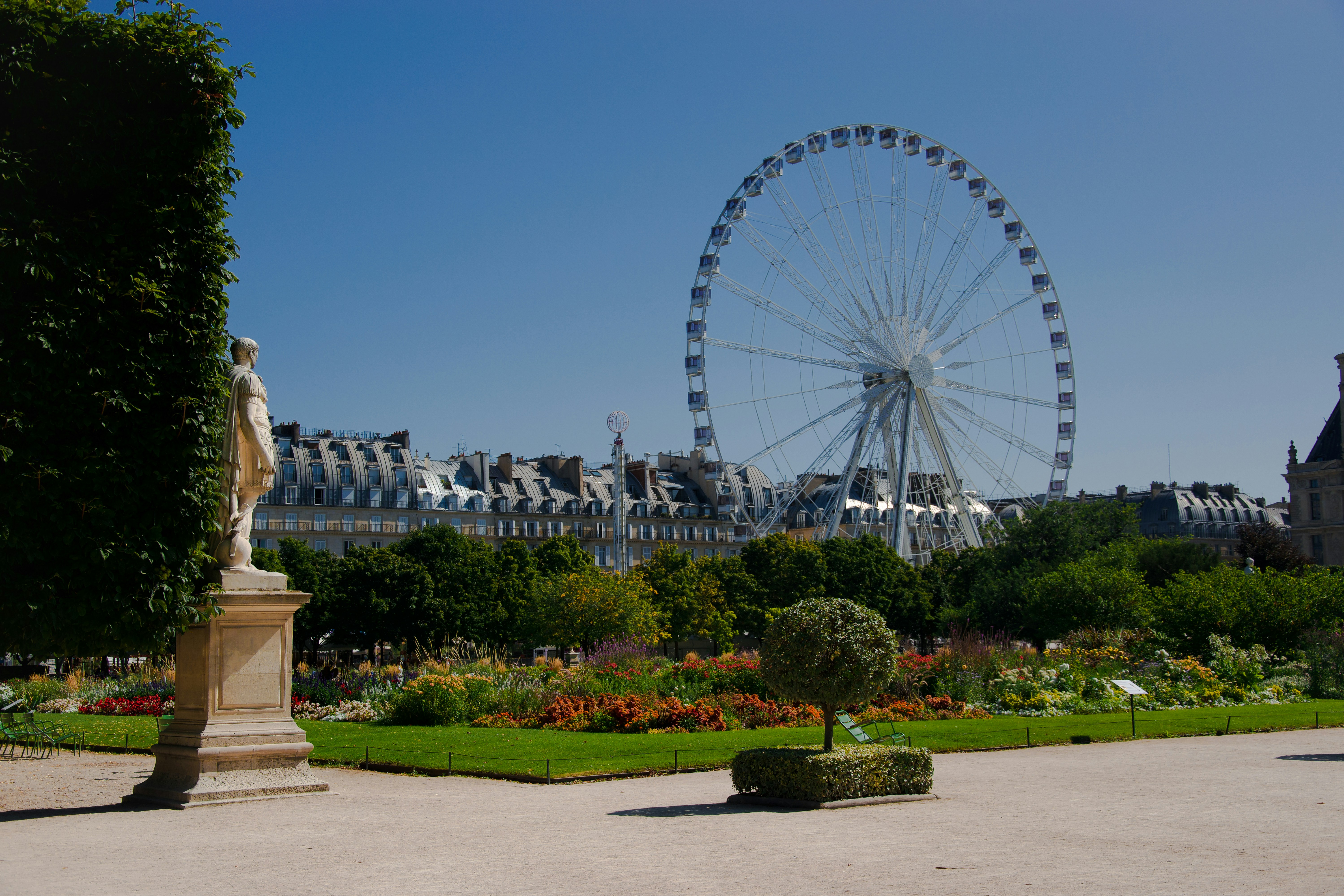 A serene garden scene featuring a stately statue and vibrant flower beds, with a large Ferris wheel in the background under a clear blue sky.