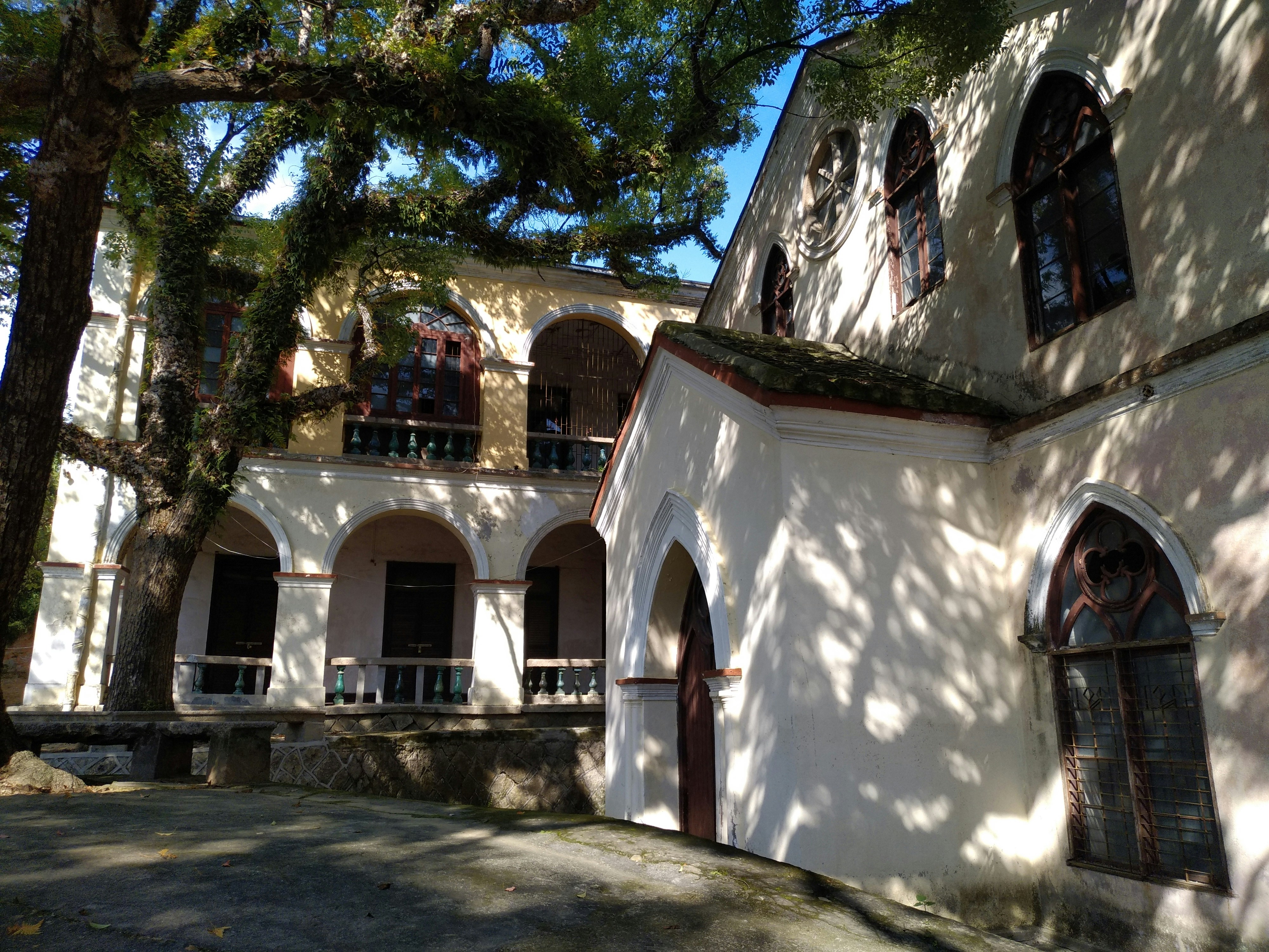 Historic white building with arched windows, partially shaded by tall trees.