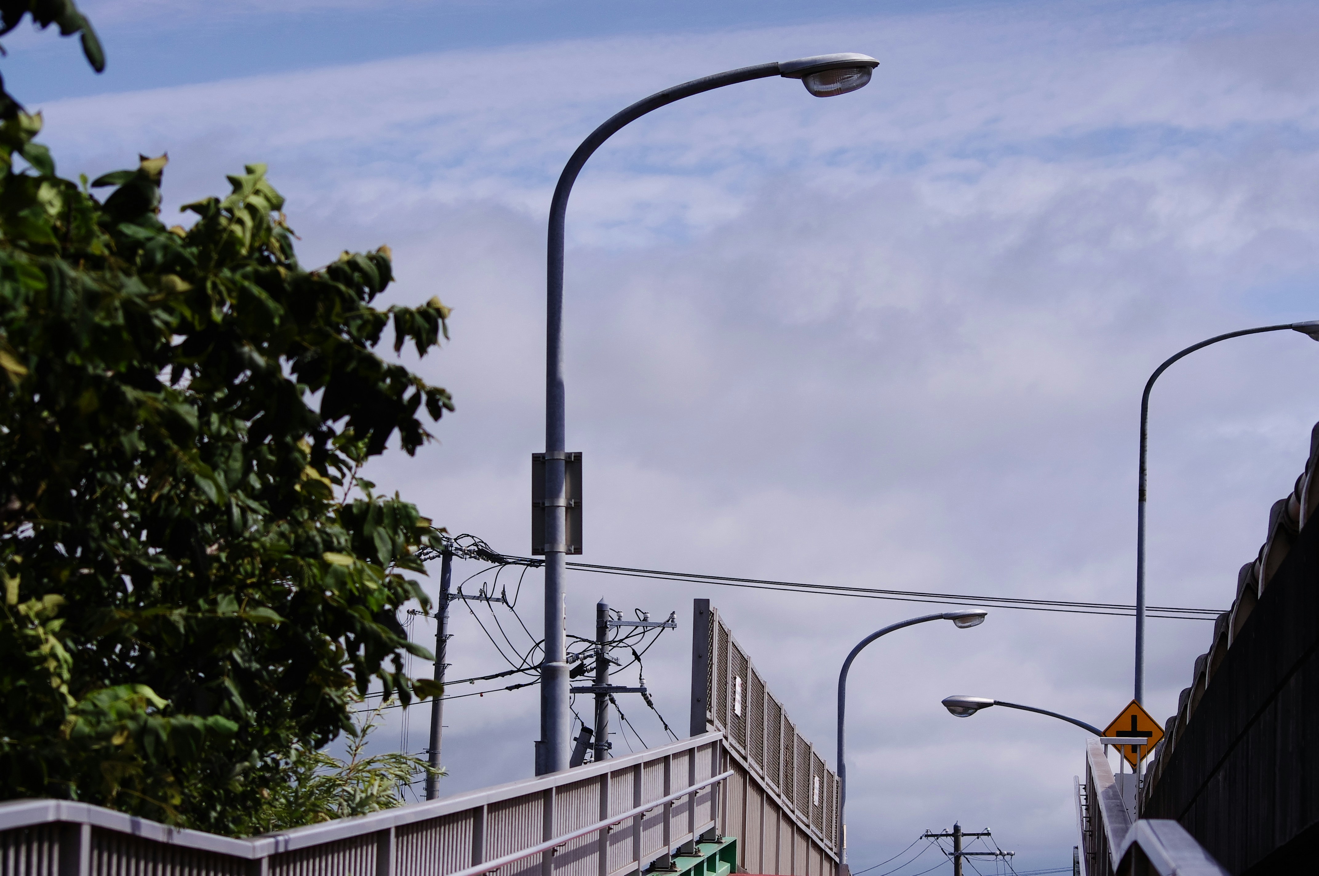 A winding pedestrian ramp leads towards a cloudy sky, framed by urban foliage and streetlights. The scene captures the essence of city life and tranquility.