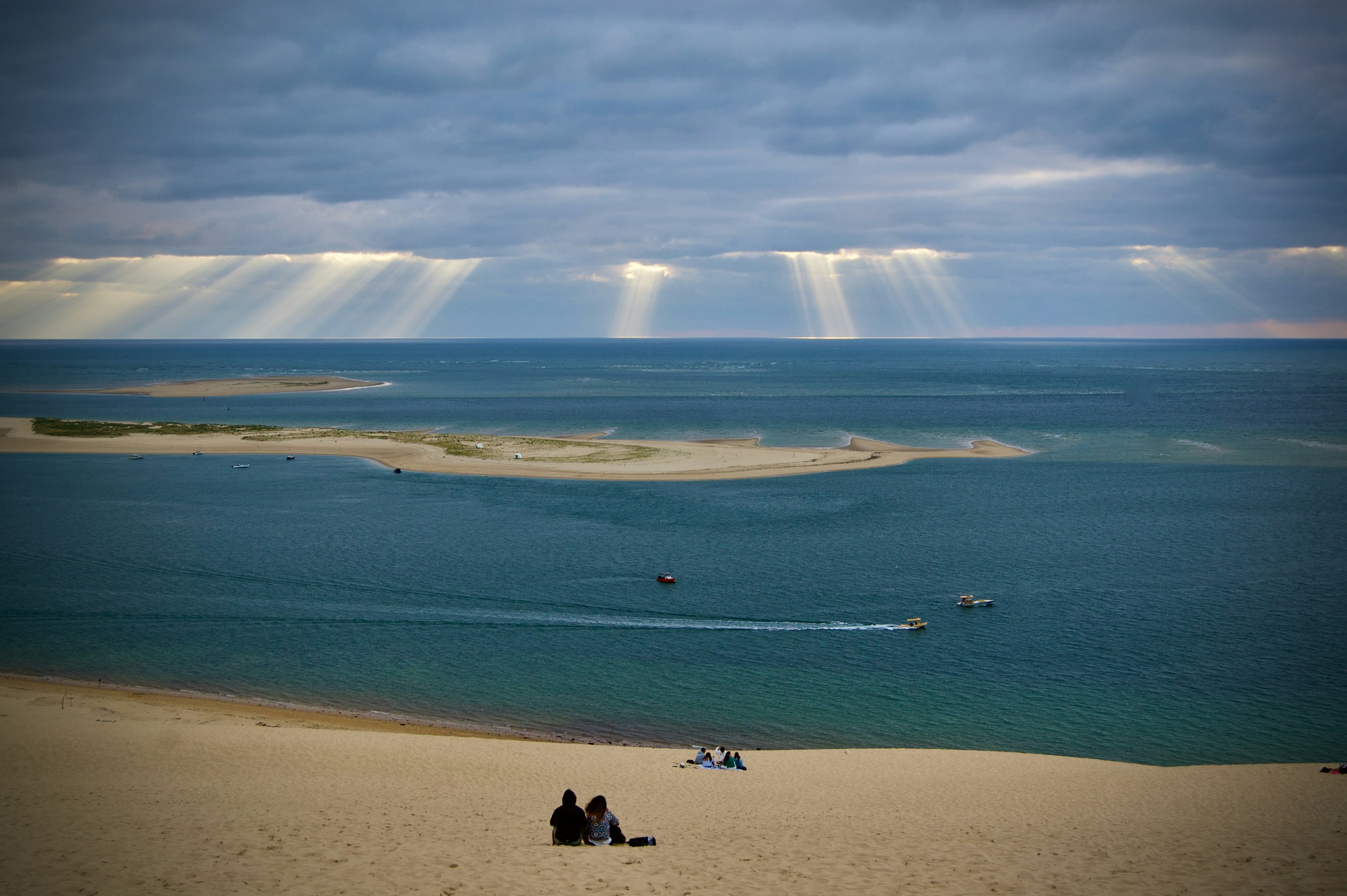 Sunbeams breaking through clouds over a vast ocean view with boats near a sandy dune.