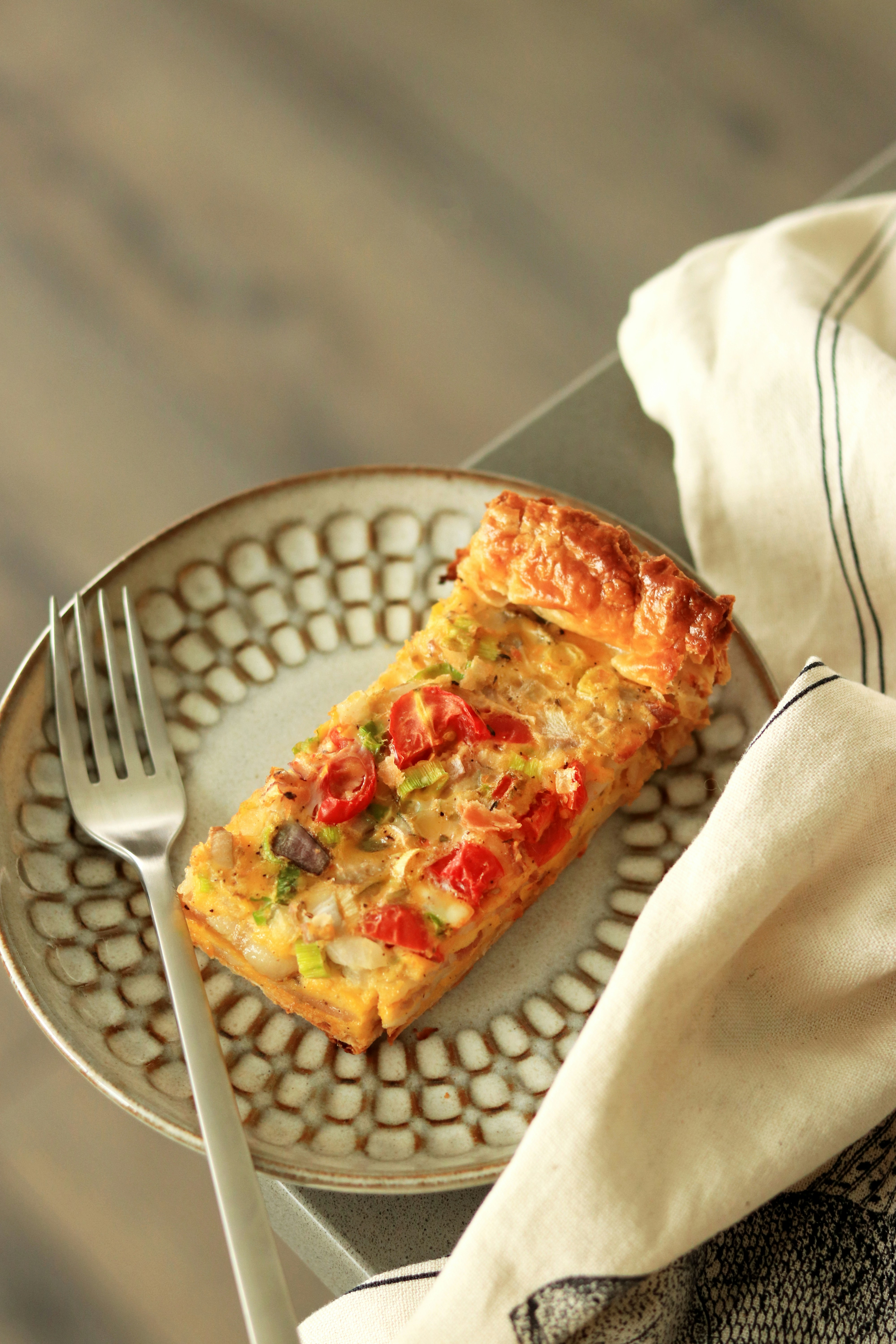 A slice of vegetable tart featuring vibrant tomatoes and leeks, elegantly presented on a textured plate with a fork beside it.