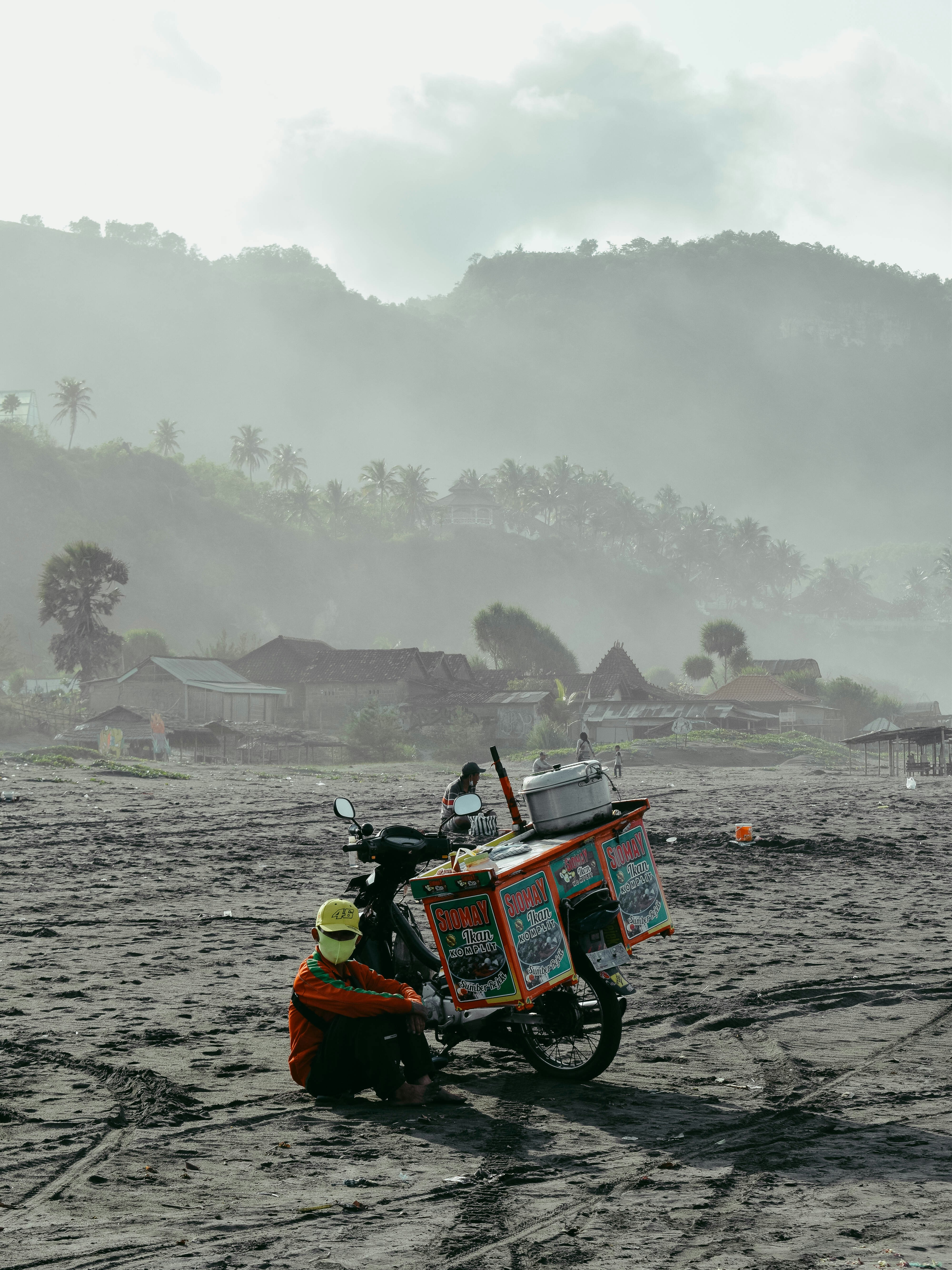 Red-jacket courier crouches beside a colorful, poster-covered delivery bike. Palm trees and fog-enshrouded hills loom in the misty riverside background.