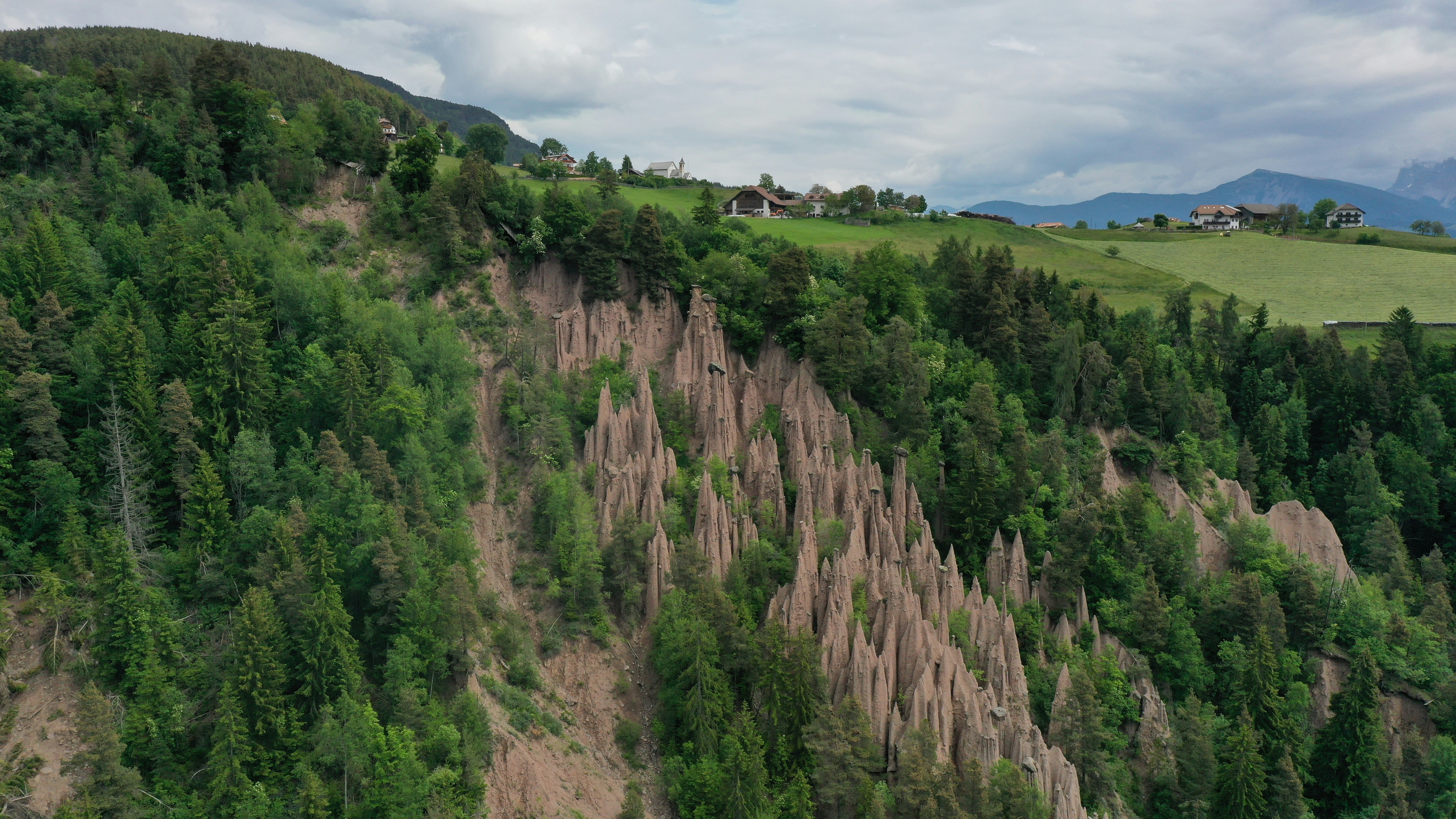 Unique geological formations rise sharply from a lush green hillside, surrounded by dense trees and distant mountains. The scene captures the harmony of nature's artistry.