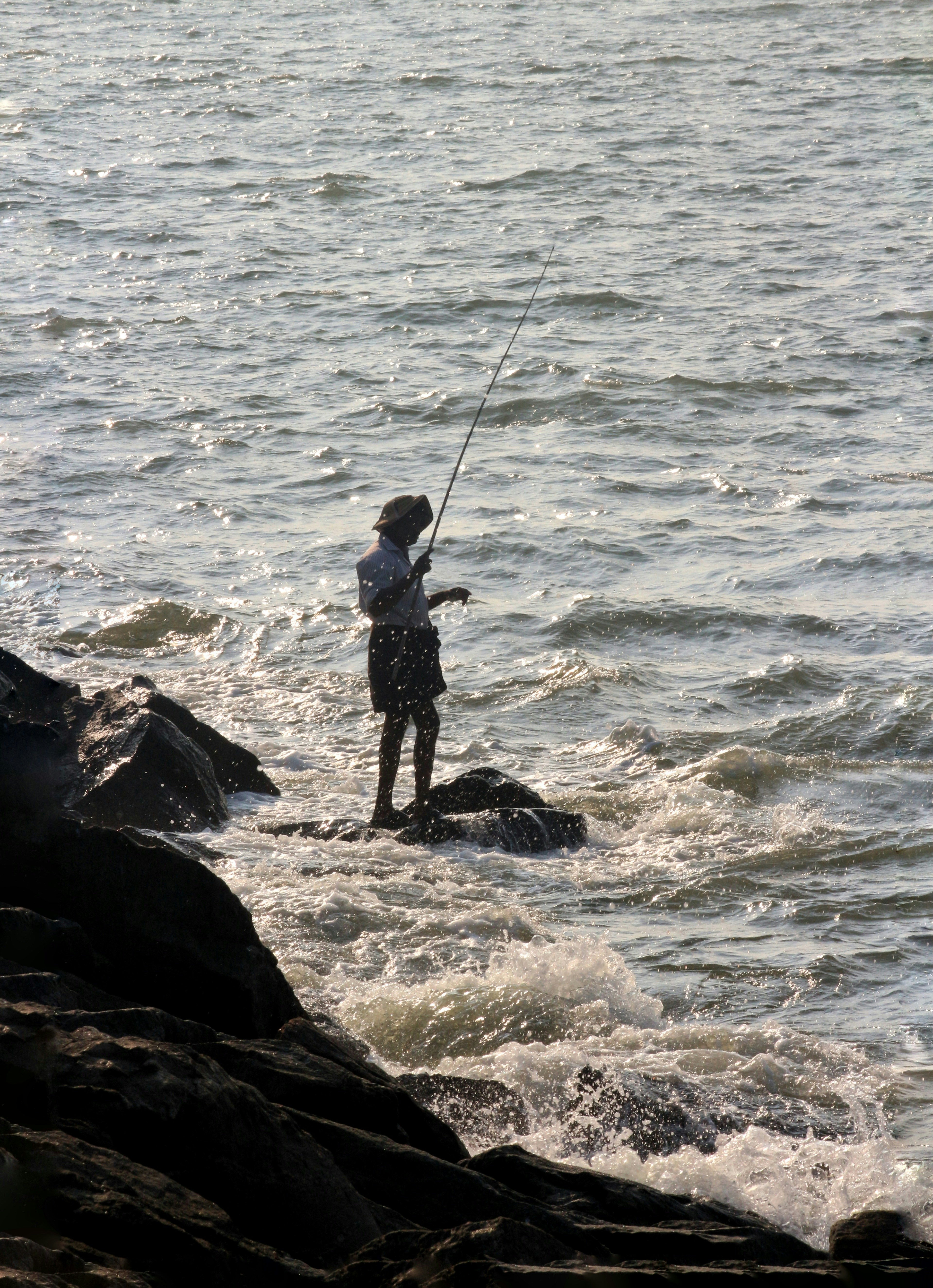 Fisherman standing on a rock, casting a line into the gently rippling sea, surrounded by splashes of water. 