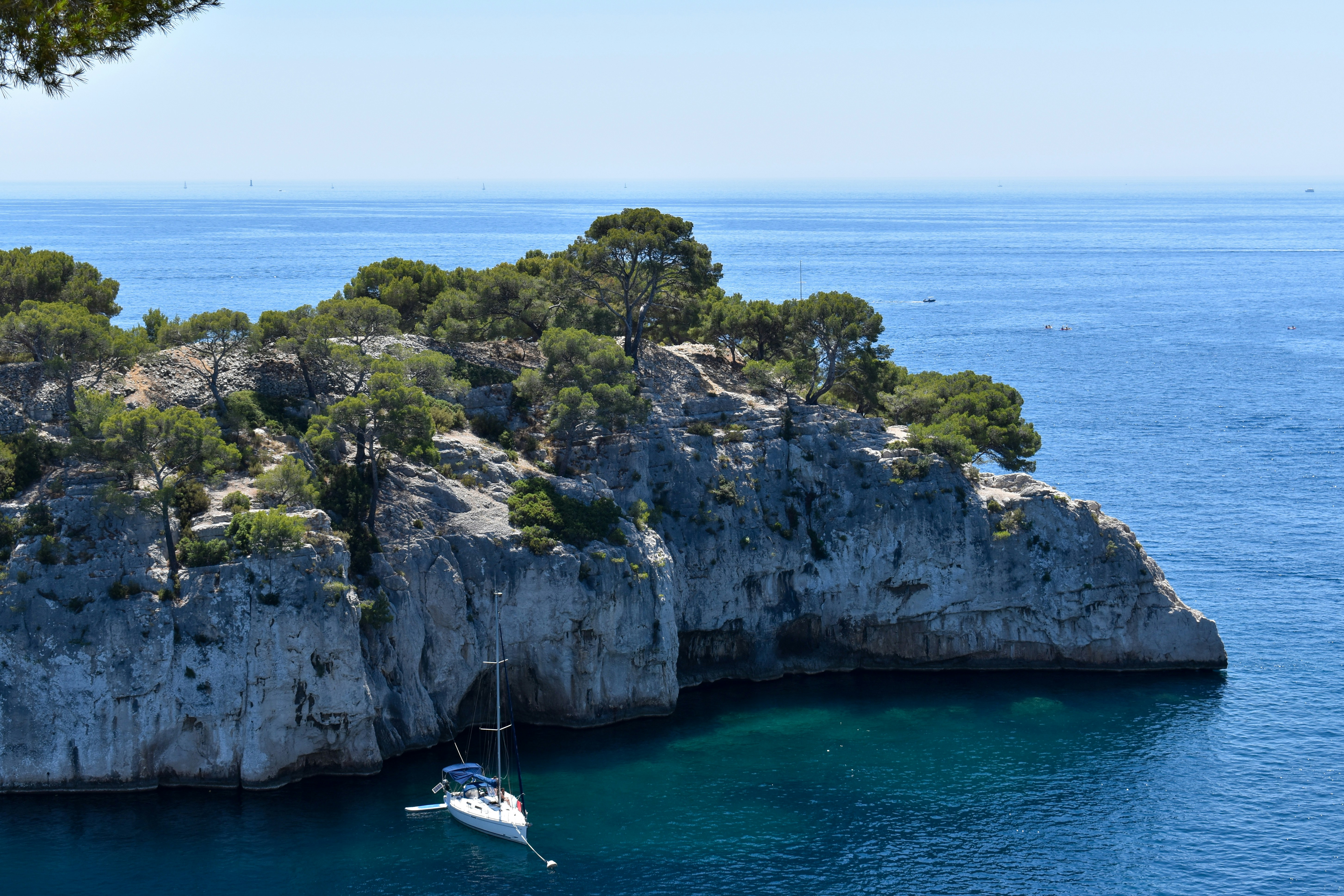 A sailboat anchored near rugged cliffs adorned with lush greenery, overlooking a tranquil blue sea. The scene captures the essence of coastal peace.