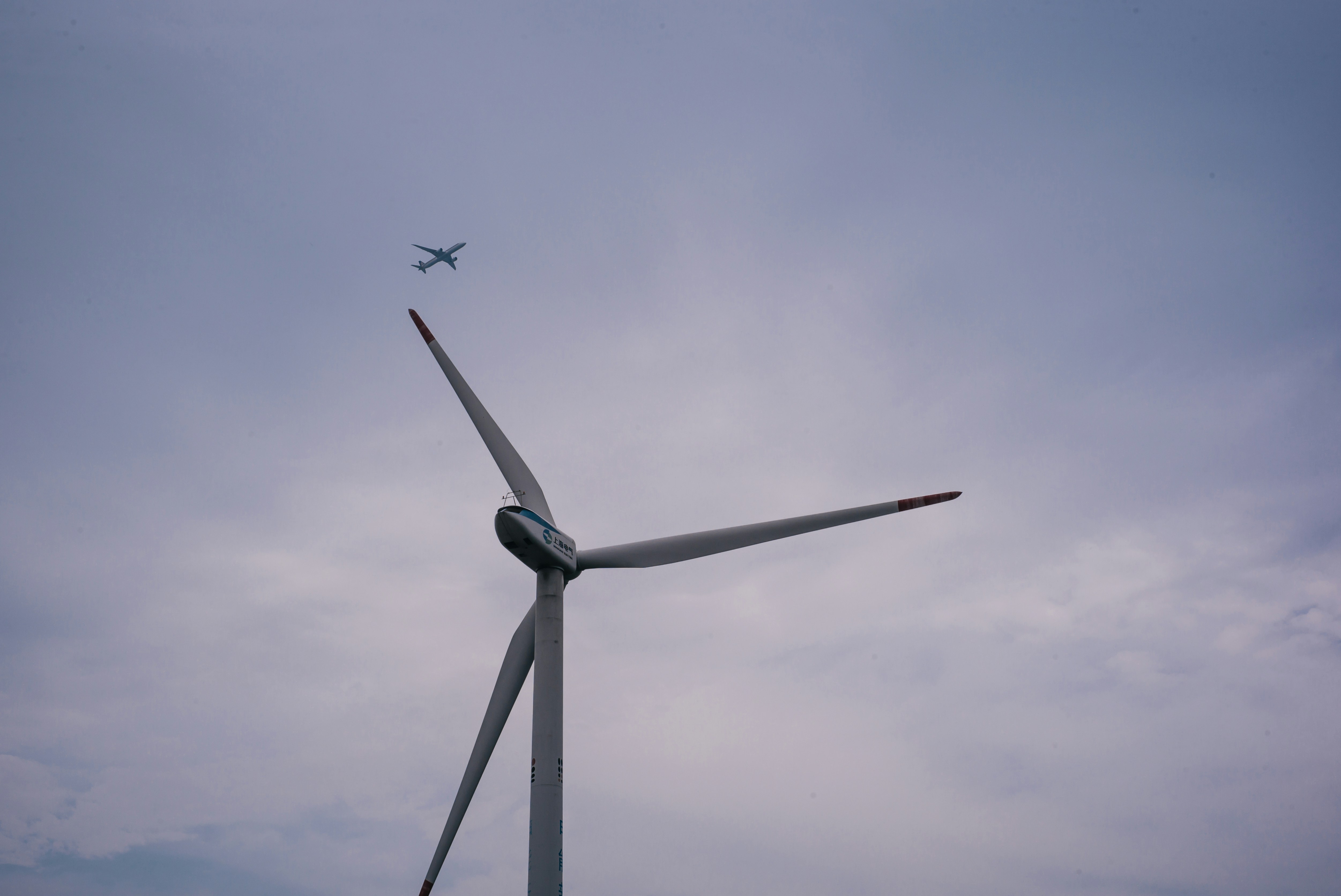 A wind turbine dominates the lower portion of the frame while a small jet passes overhead against a cloudy sky. The scene emphasizes scale and industrial form.