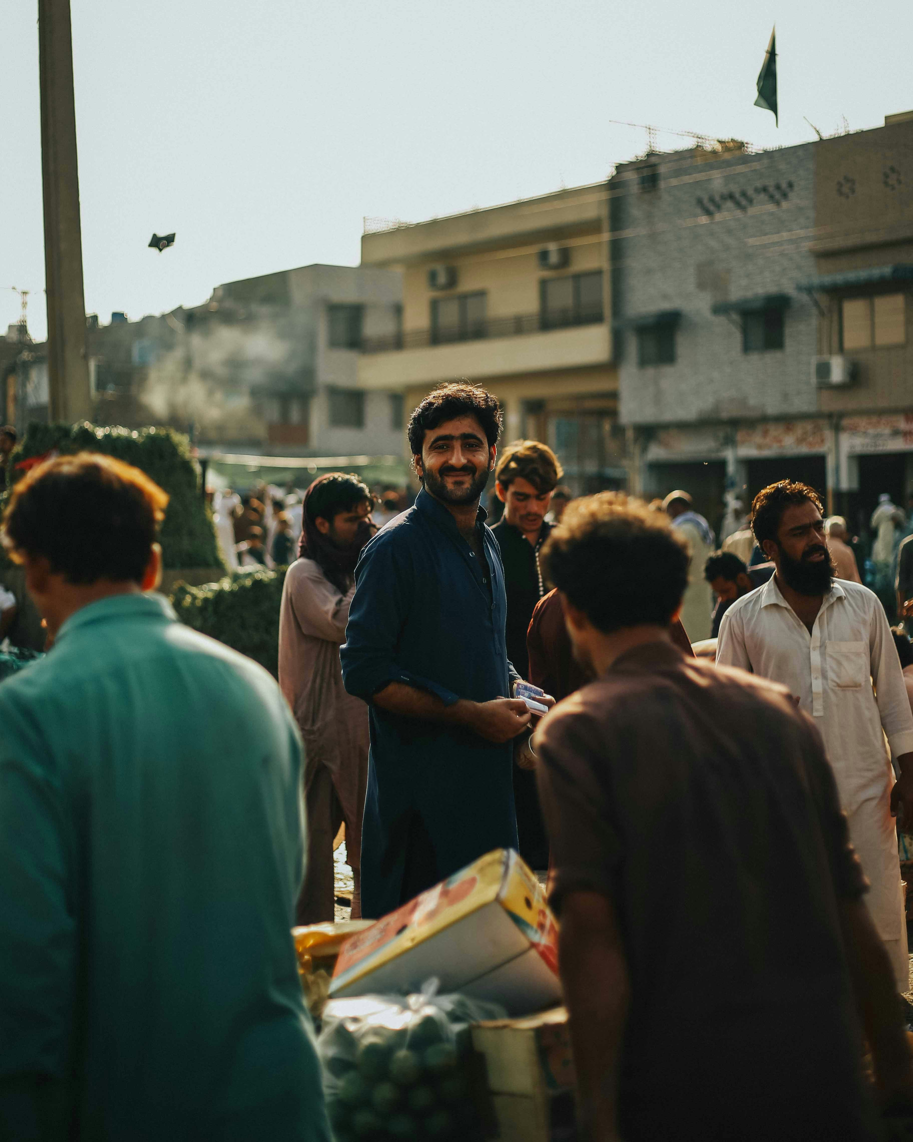 A bustling market scene with a young man smiling amidst a crowd, surrounded by vendors and produce. The vibrant atmosphere captures the essence of local commerce.