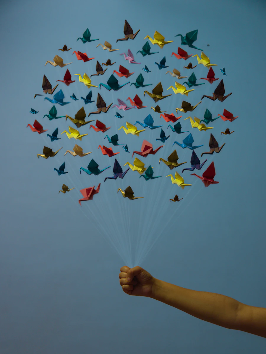 A clenched hand at the bottom of the frame sends a swarm of colorful origami birds upward against a plain blue sky-like background.