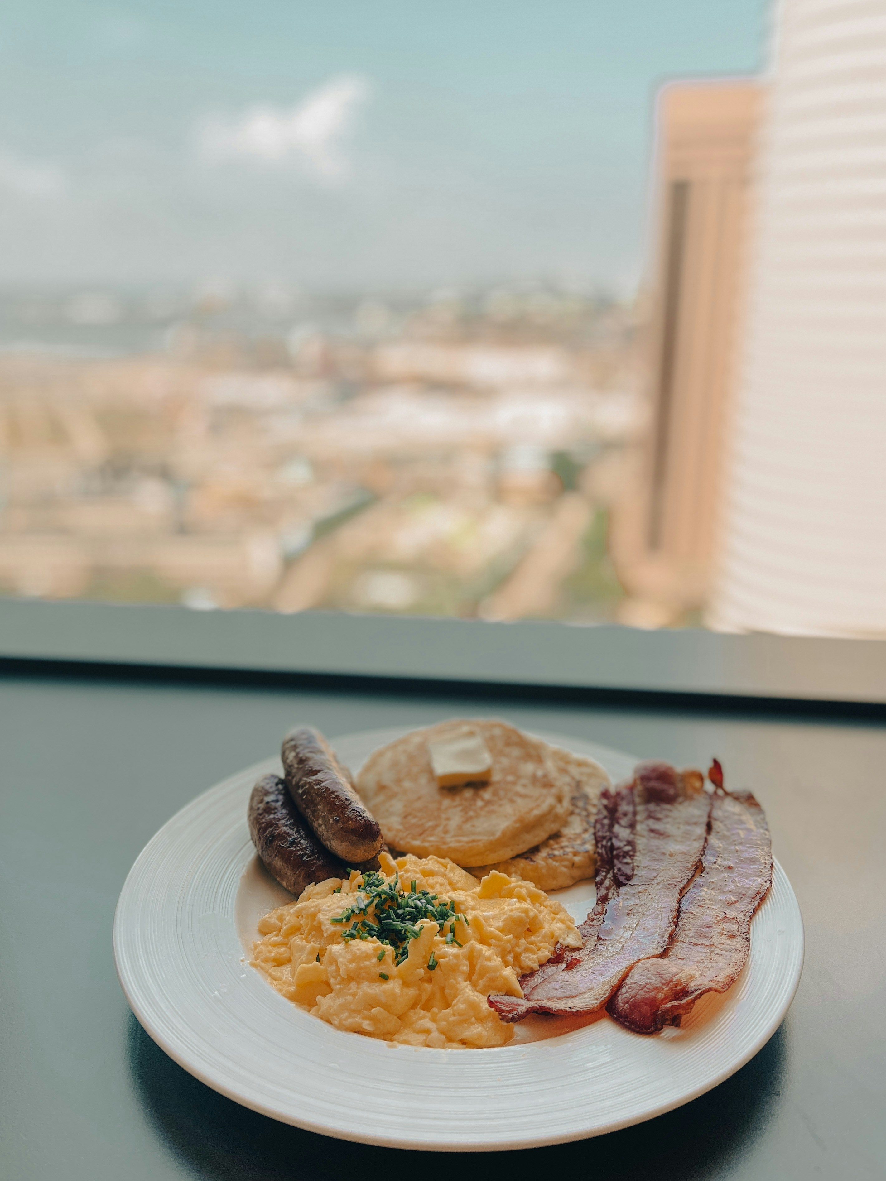 Window-side Breakfast Mosaic: Creamy Eggs, Crispy Bacon, and Pancakes with a City View