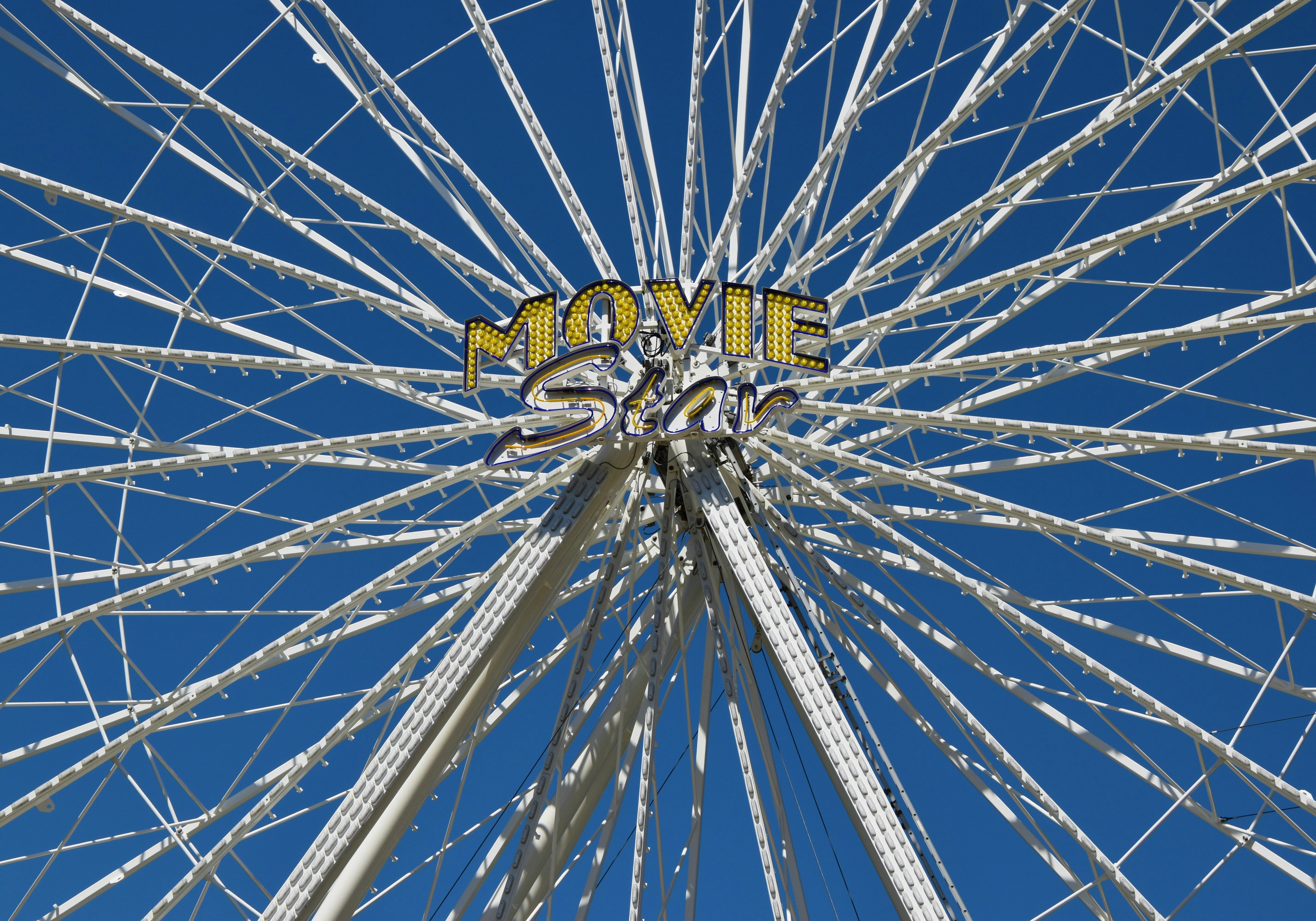 The iconic Movie Star Ferris Wheel showcases its vibrant lettering against a clear blue sky, emphasizing its playful design and attraction. The intricate web of spokes adds depth and interest.