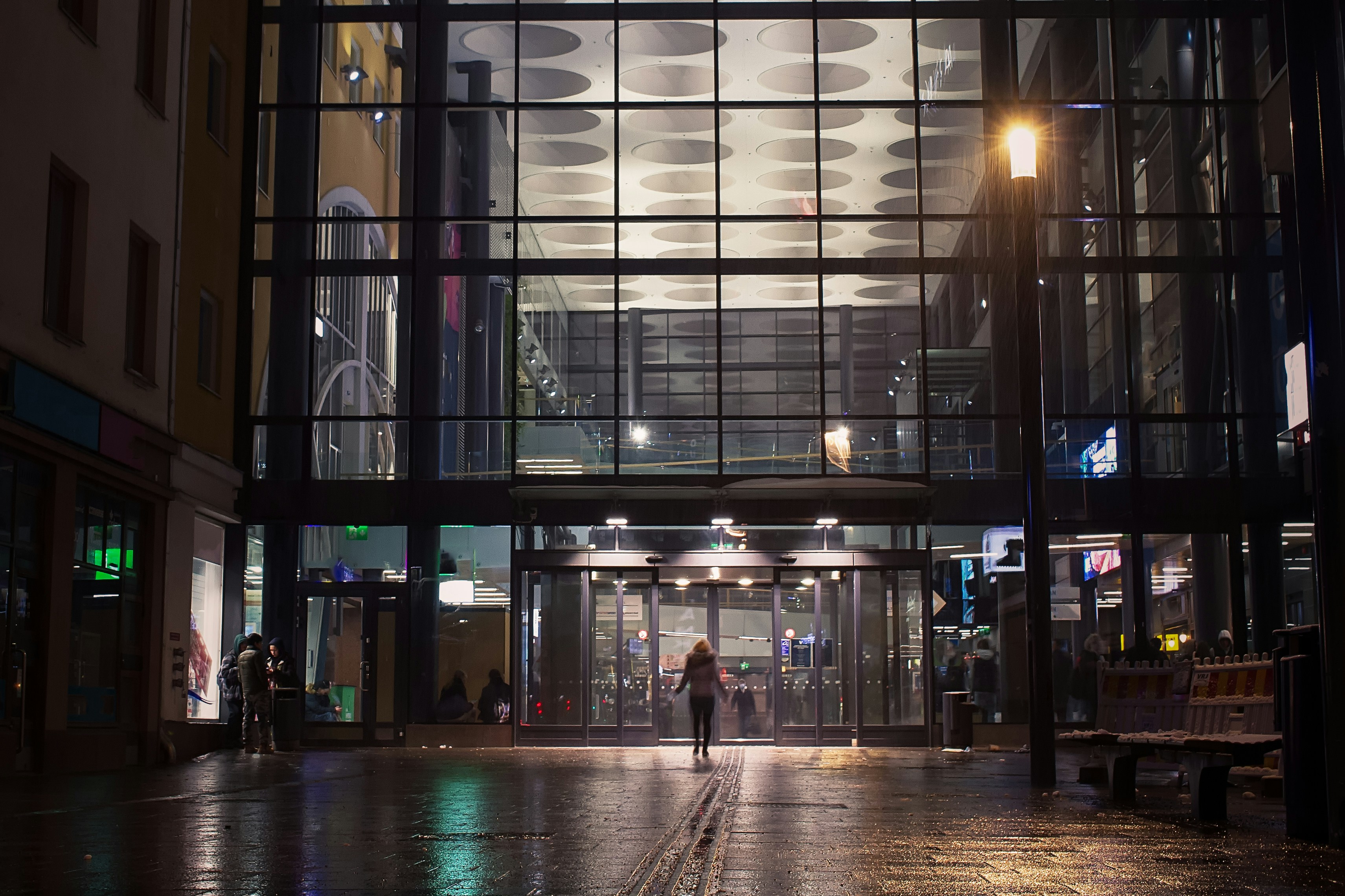 Person entering a glass-fronted shopping center at night, with illuminated round ceiling patterns contrasting rectangular facade elements.