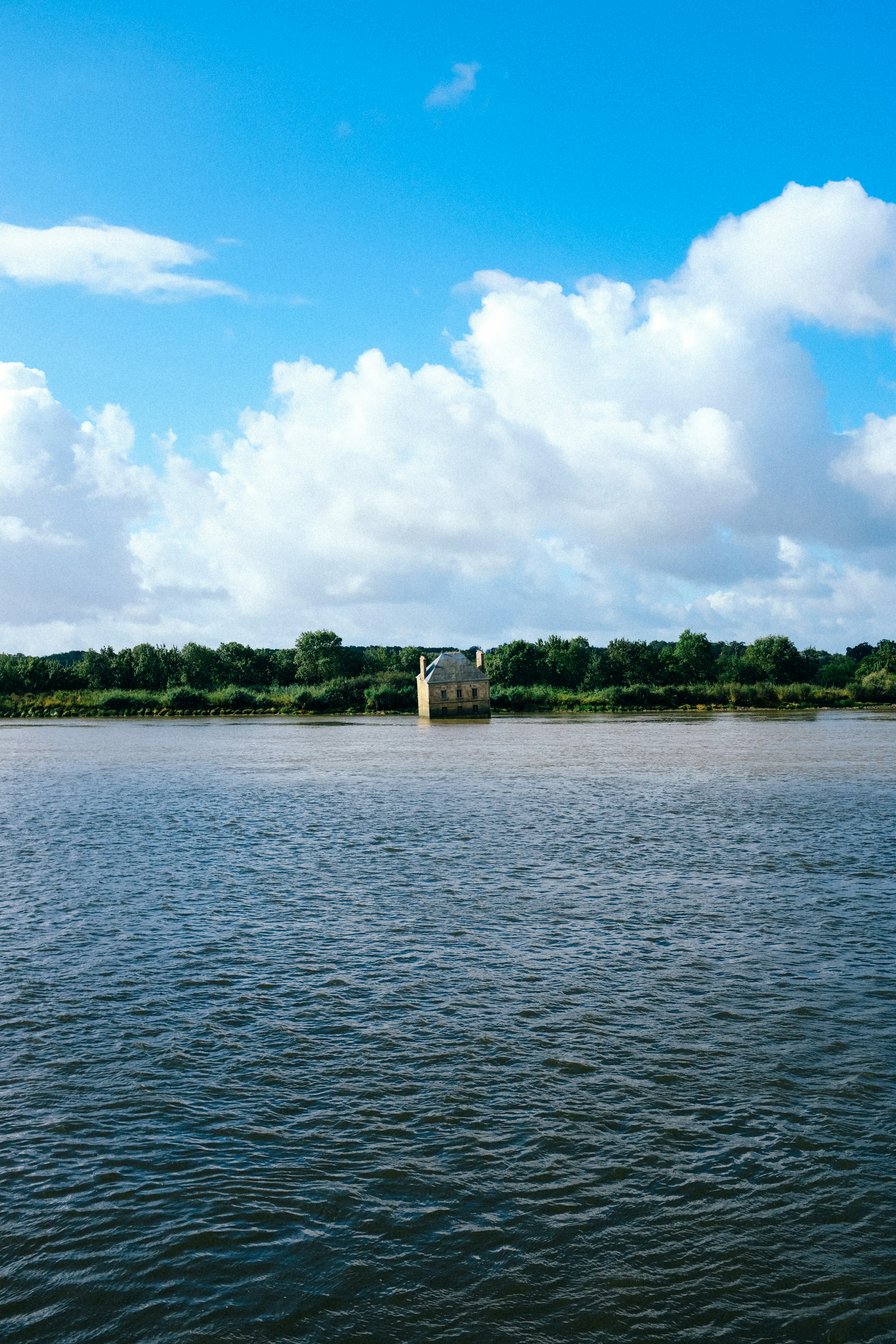 A quaint building stands alone by the riverbank, surrounded by lush greenery and under a vibrant sky filled with clouds.