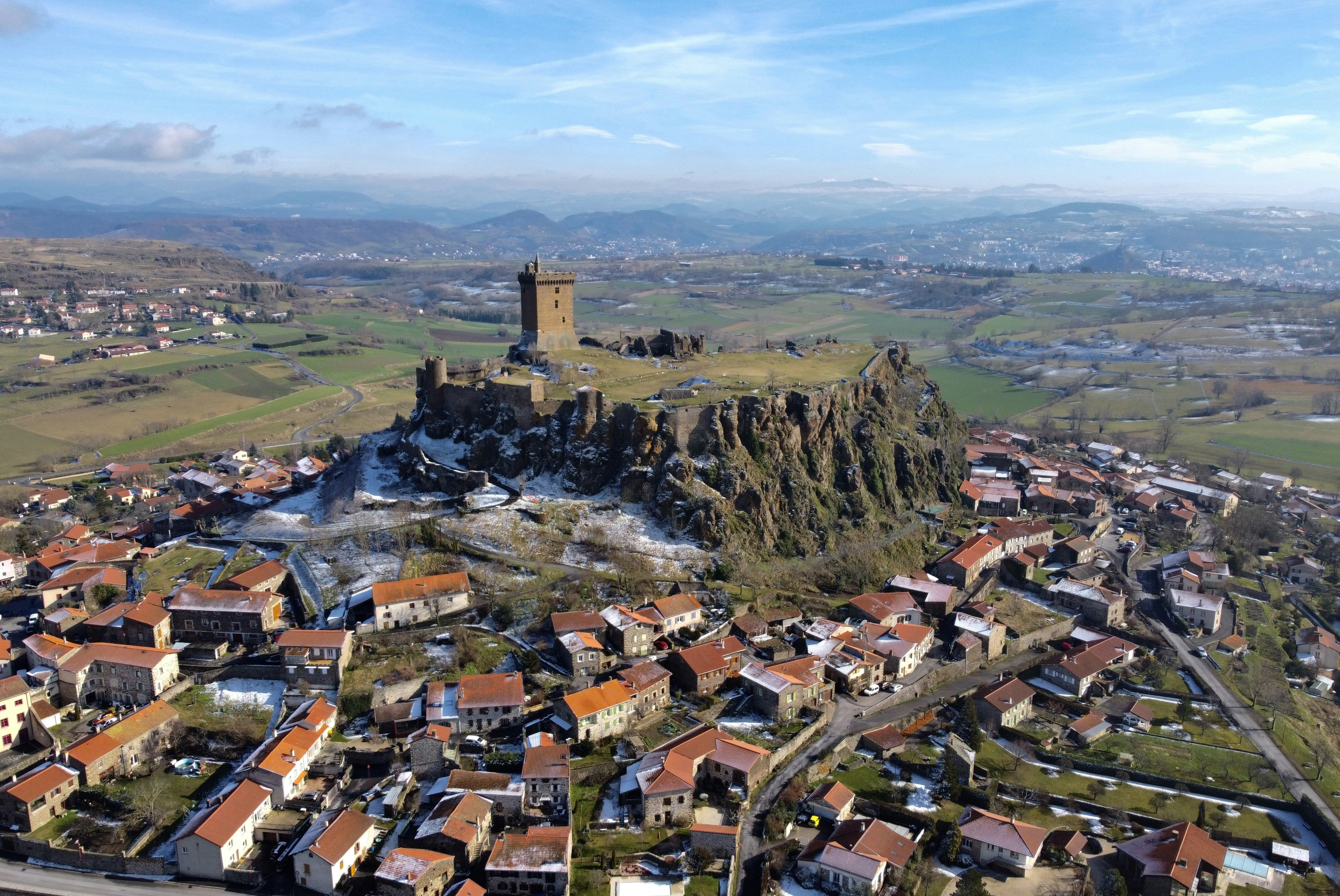 Medieval fortress atop a rocky hill surrounded by a quaint village and sprawling countryside under a clear blue sky.