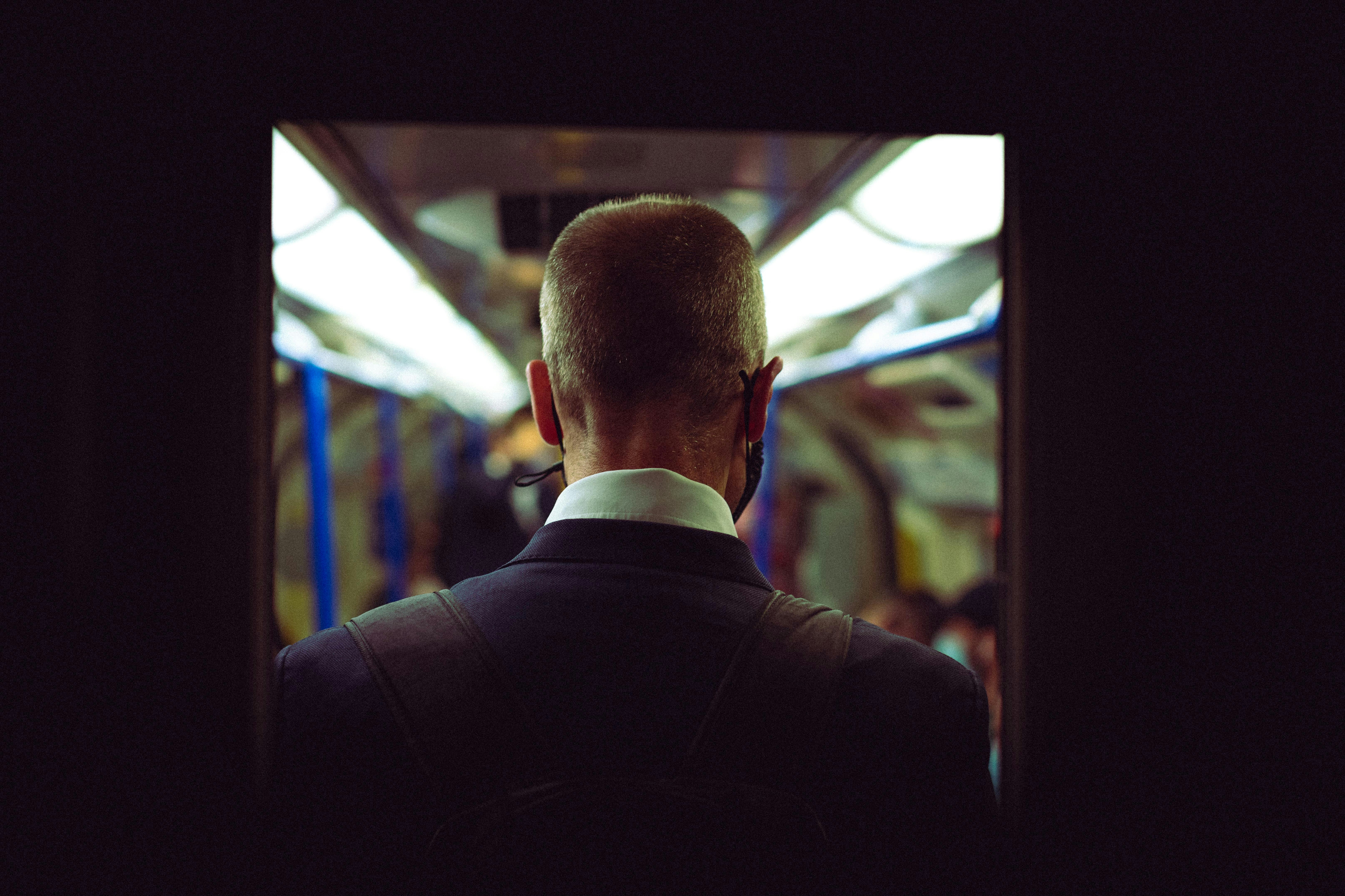 This evocative image captures the back of a solitary commuter standing in a dimly lit subway car, surrounded by blurred passengers. The composition is framed by the train's doorway, highlighting the contrast between the sharp silhouette of the individual and the soft, ambient lighting of the interior. The muted colors and shadows create a contemplative atmosphere, drawing attention to the feeling of isolation amidst the bustling crowd.