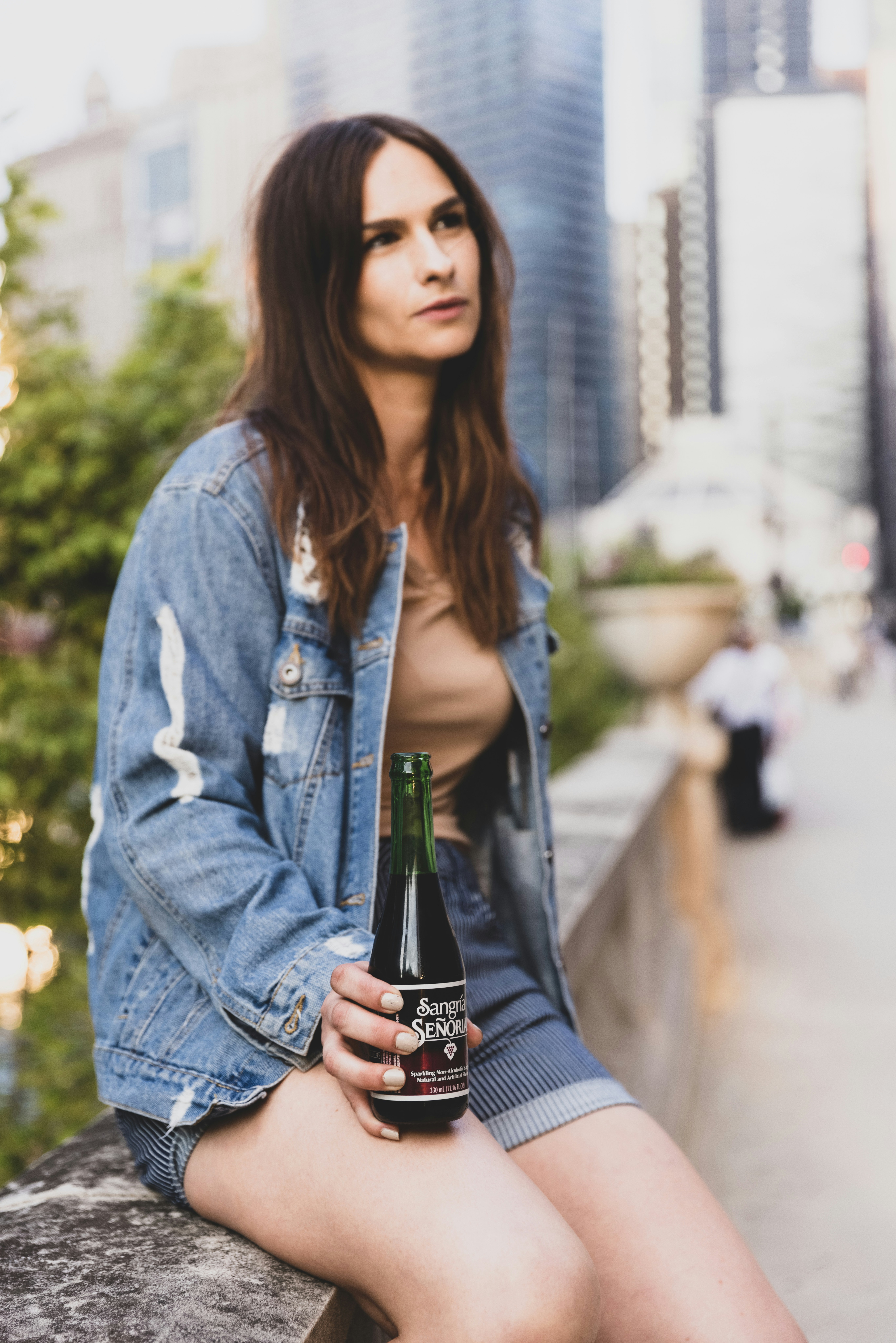 A woman in a denim jacket sits casually on a ledge, holding a bottle while gazing thoughtfully into the distance amidst a city backdrop.