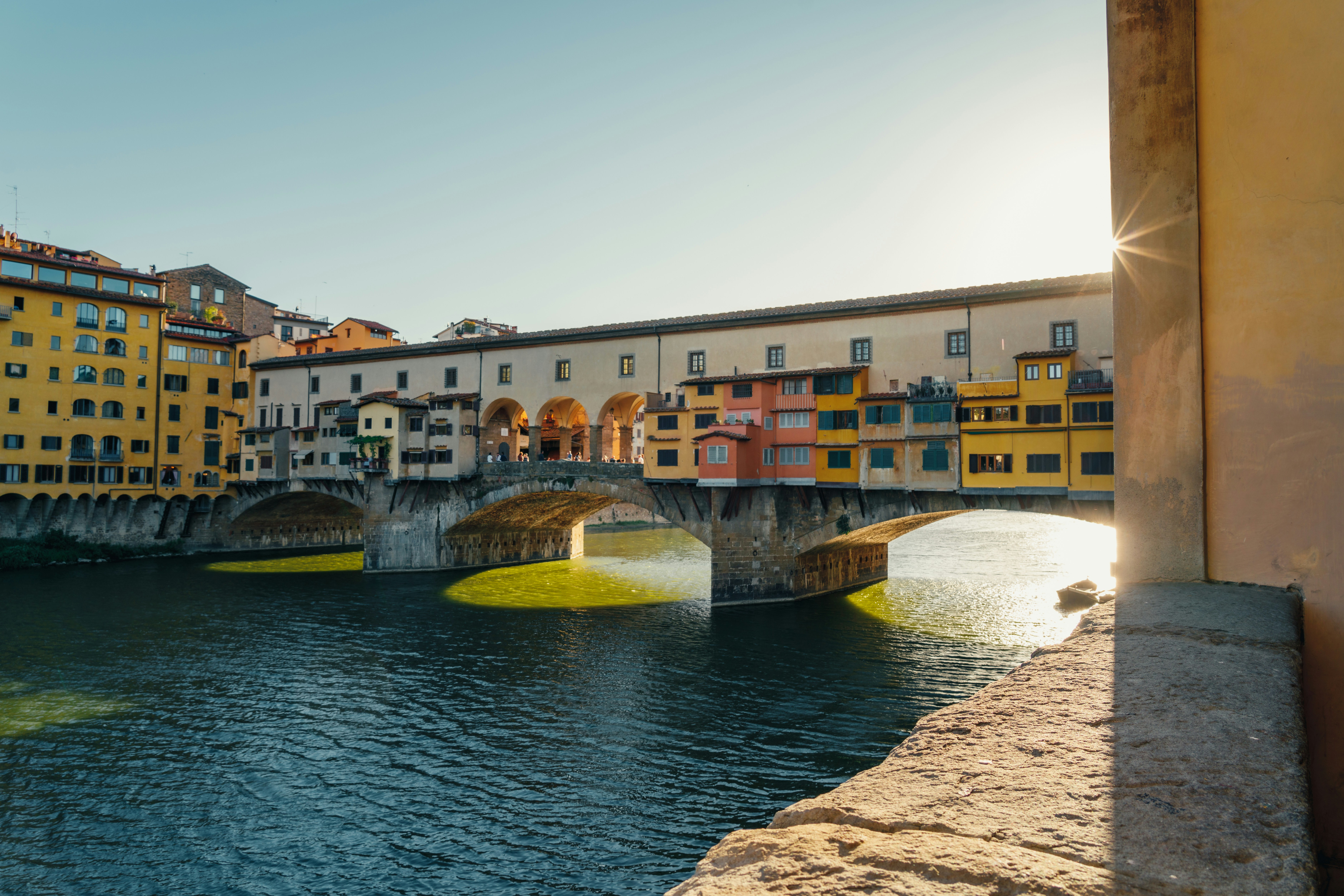 The Ponte Vecchio bridge arches gracefully over the Arno River, framed by colorful buildings reflecting the warm glow of sunset.