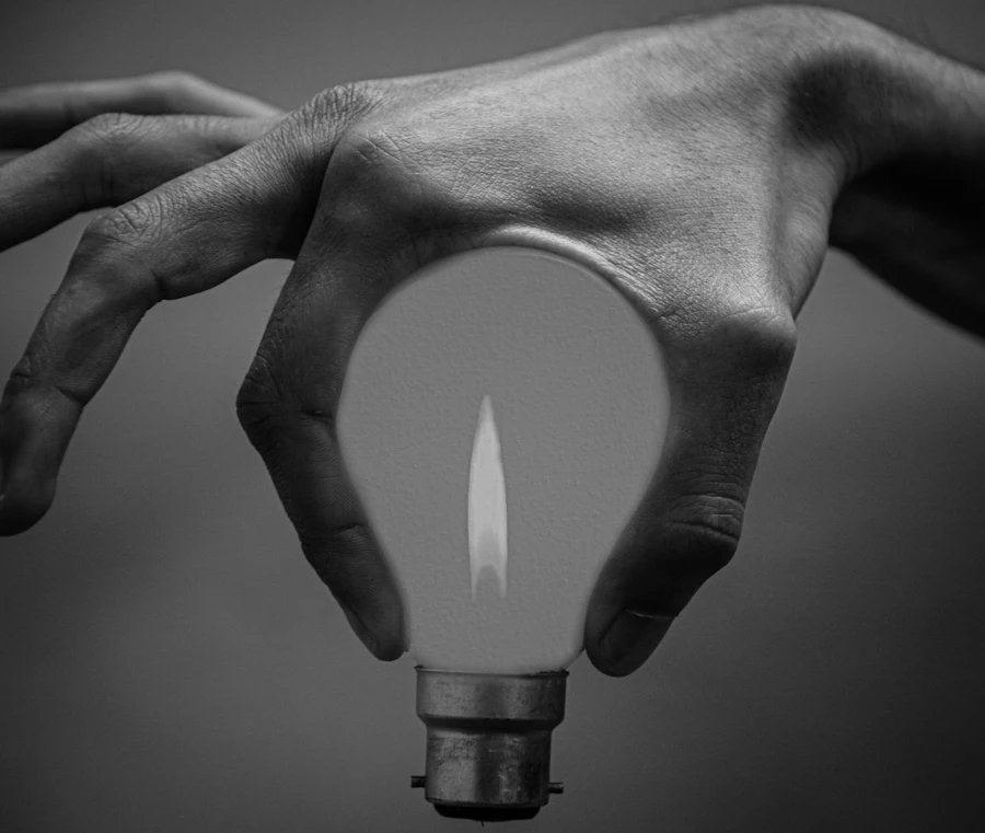 Monochrome close-up of hands cupping a glowing light bulb against a dark background. The bulb’s filament and the hands’ texture create a dramatic, sculptural composition.