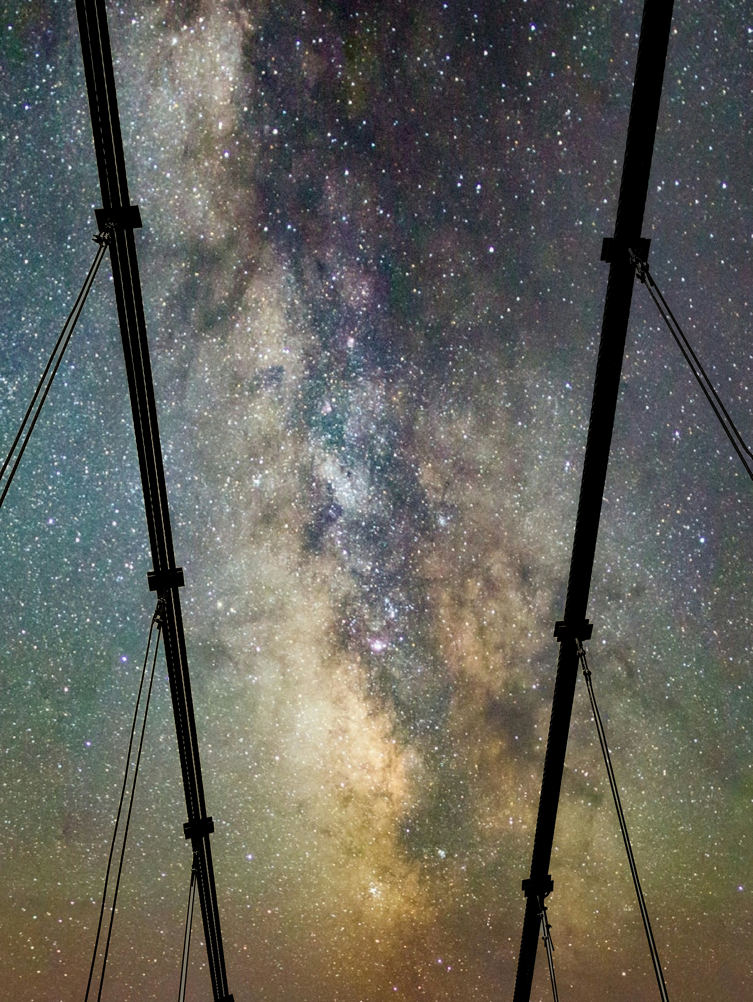 Night sky photograph of the Milky Way's bright core, framed by tall suspension cables.