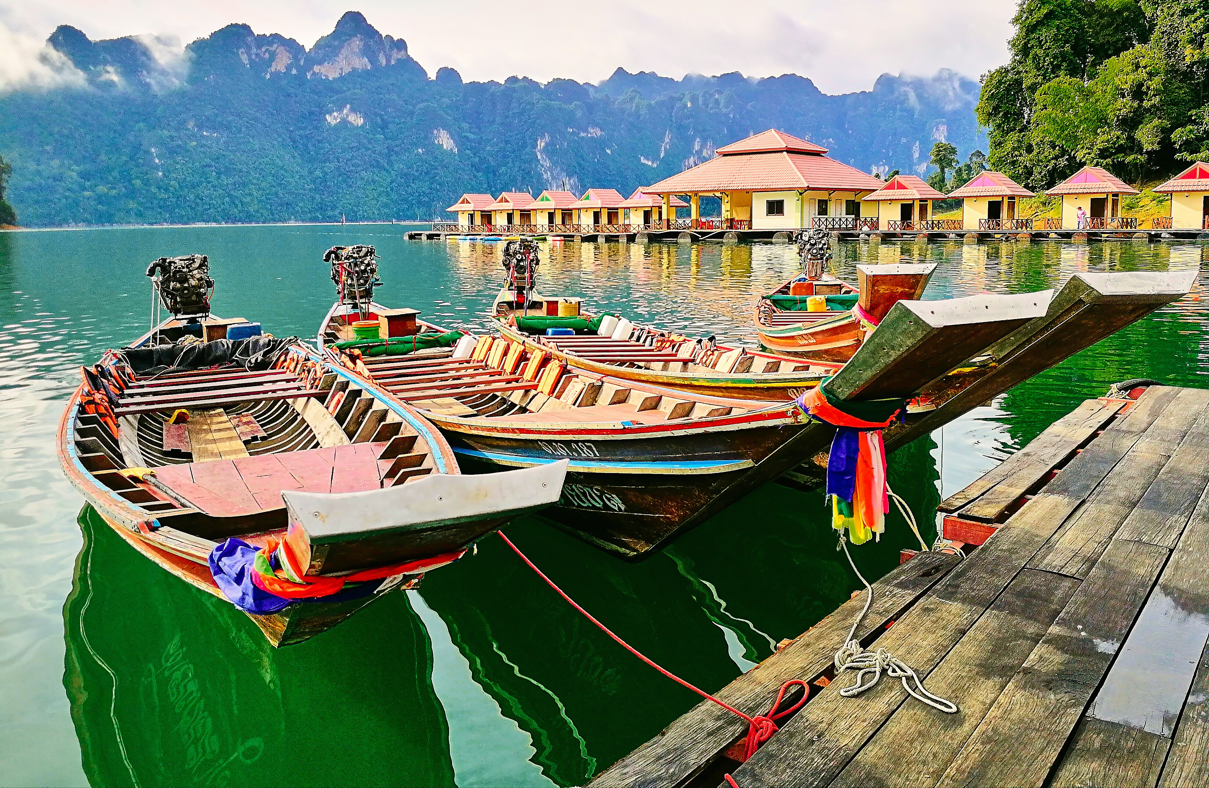 Traditional longtail boats docked on a serene lake with floating houses and mountains in the background.