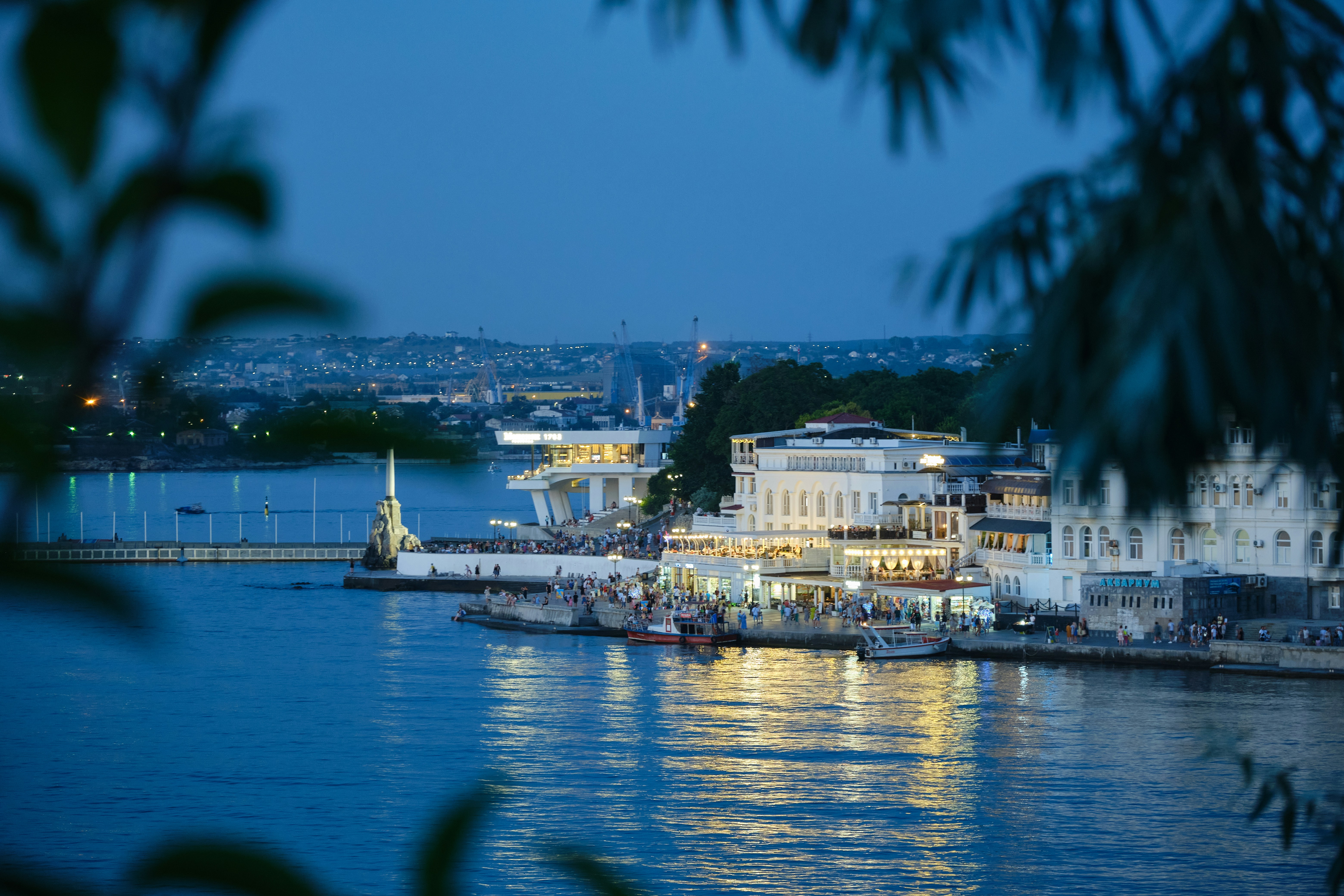 A serene waterfront scene at twilight, showcasing illuminated buildings along the shore and a gentle reflection in the water.