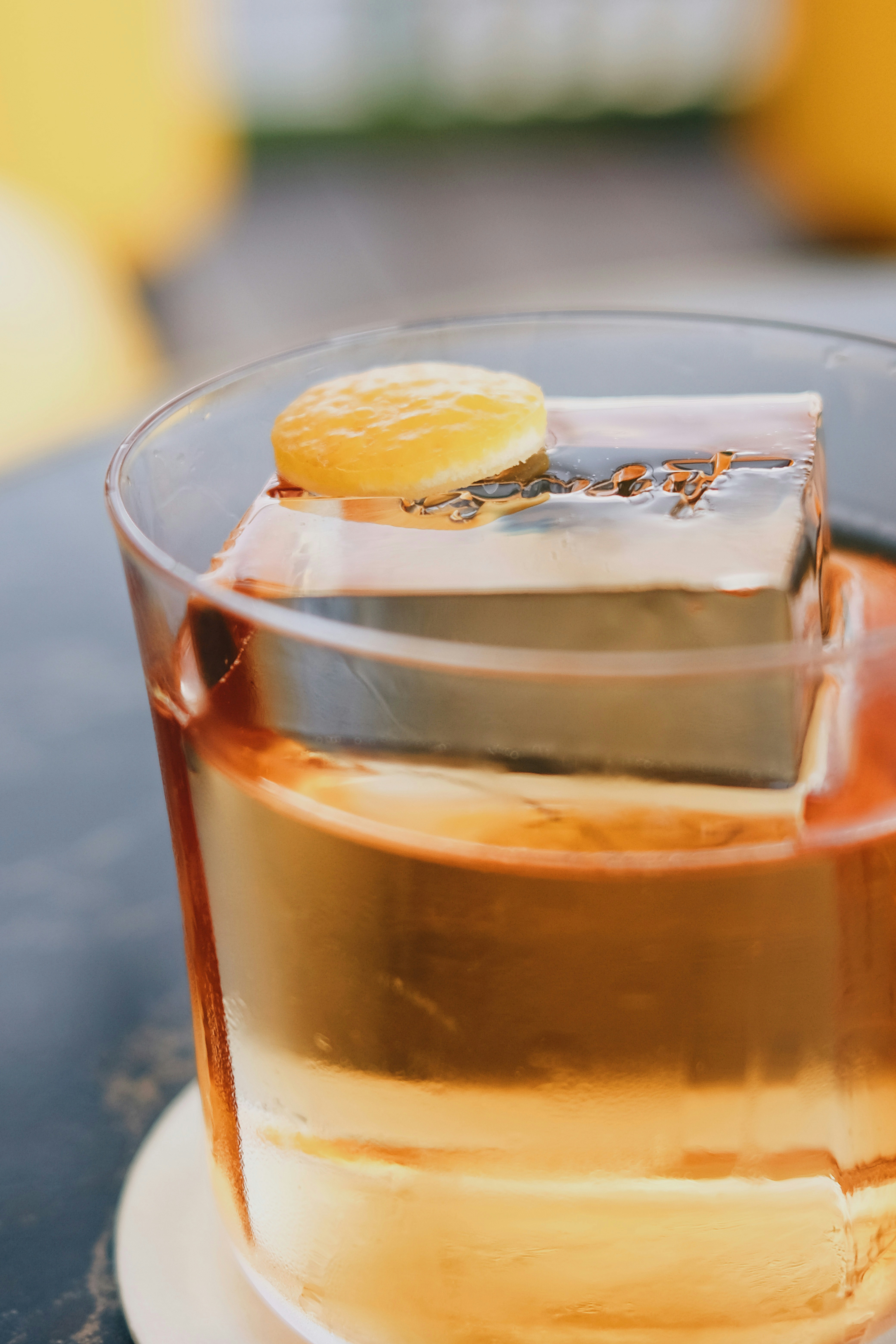 A close-up of a cocktail featuring a large ice cube topped with a slice of citrus, showcasing the drink's rich amber hues and clarity. The background hints at a vibrant setting.
