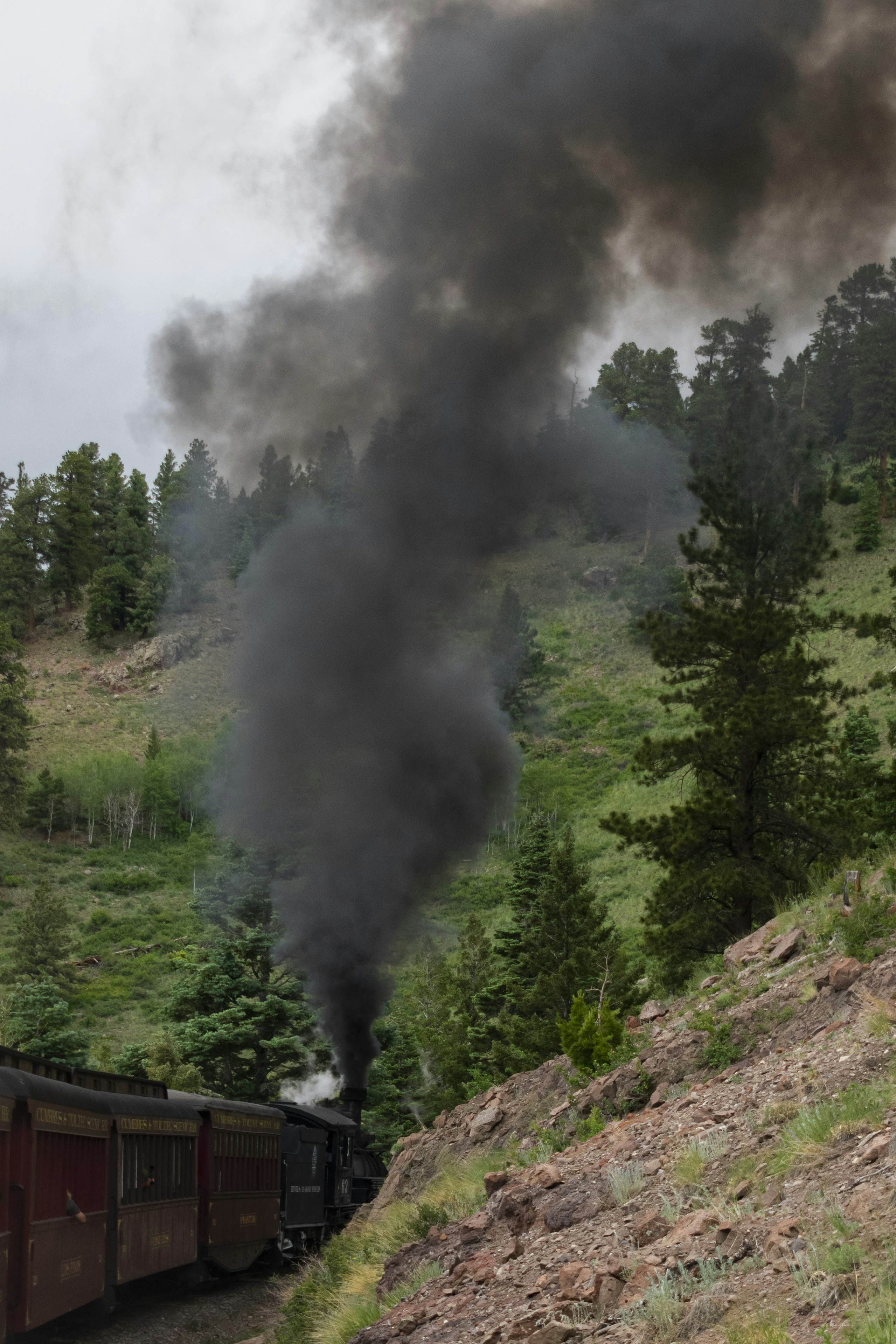 Vintage steam locomotive billowing dark smoke as it traverses a lush, mountainous landscape.