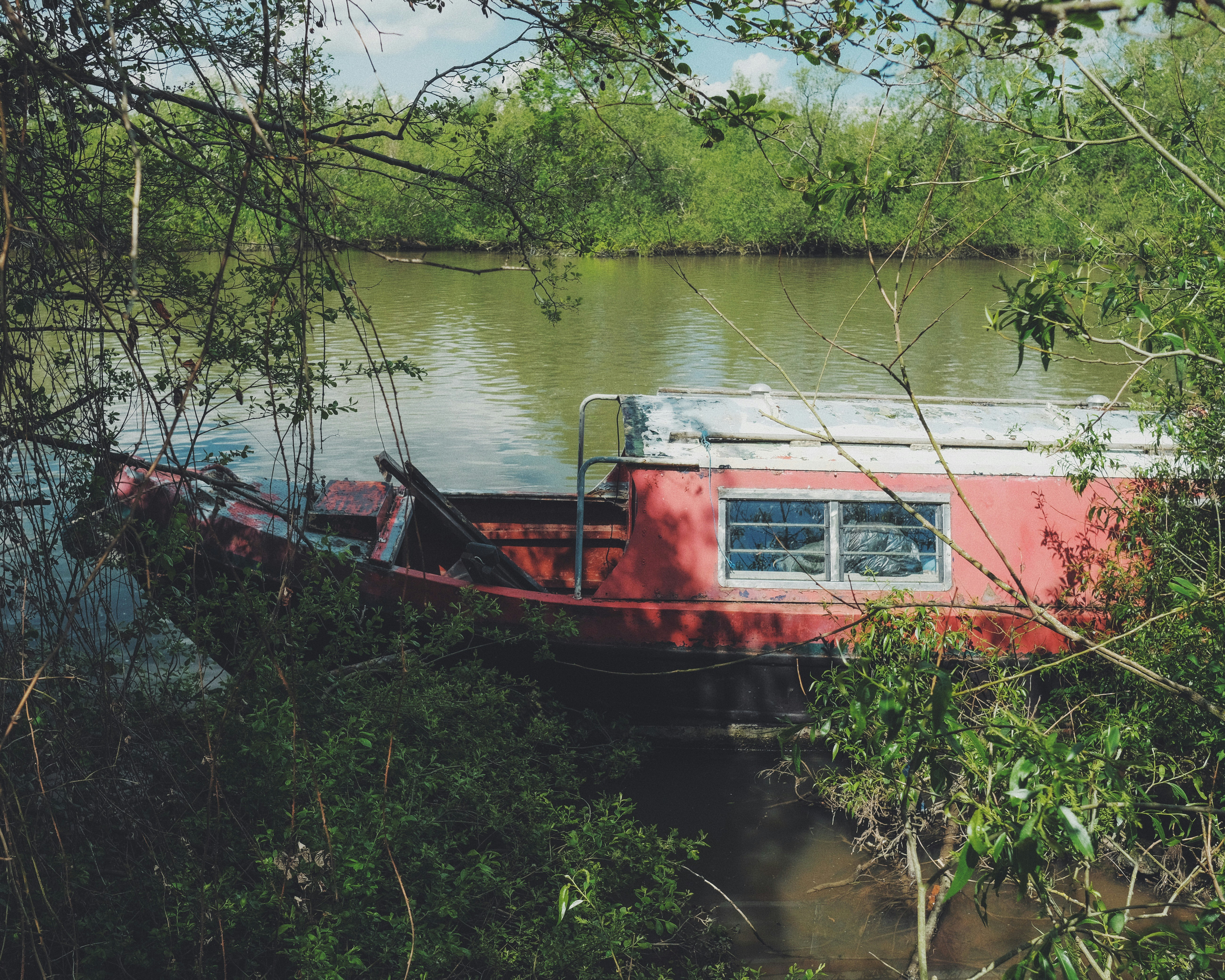 Abandoned red boat partially submerged in a tranquil river, surrounded by lush greenery and reflections on the water's surface.