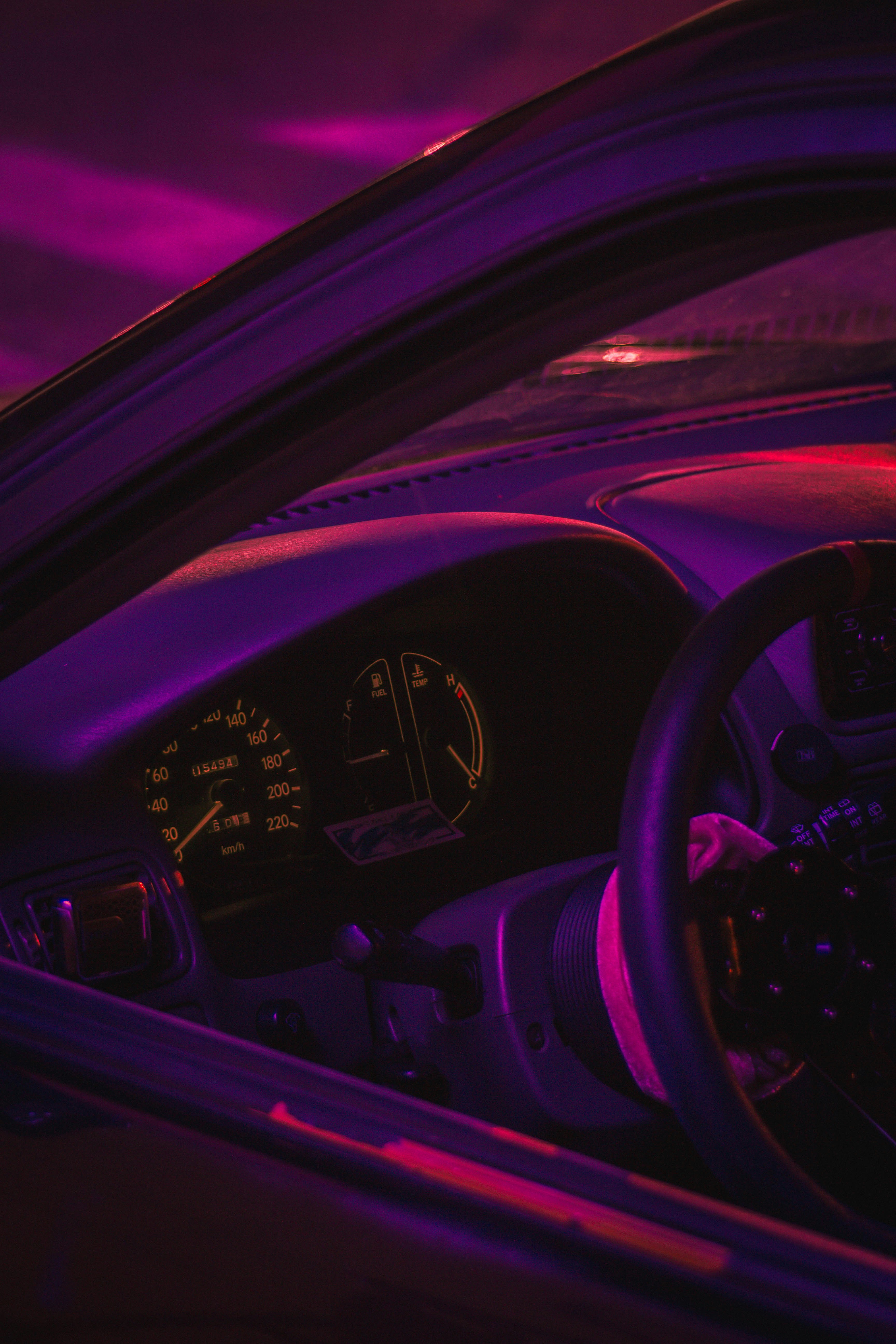 Interior view of a car dashboard illuminated by vibrant purple and pink lights, showcasing the speedometer and steering wheel.