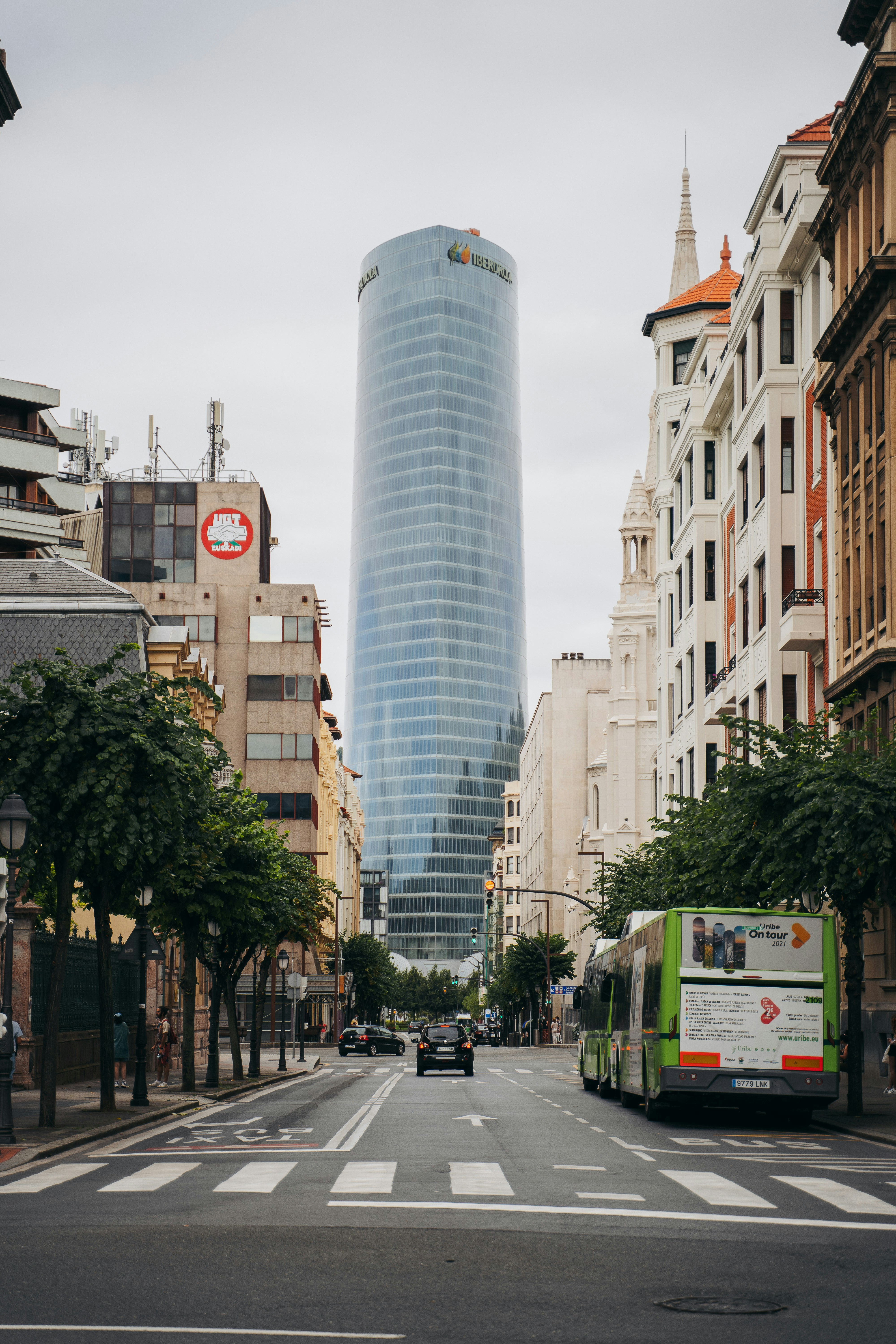 A modern glass skyscraper rises majestically amidst historic buildings, framed by leafy trees lining the bustling street below.