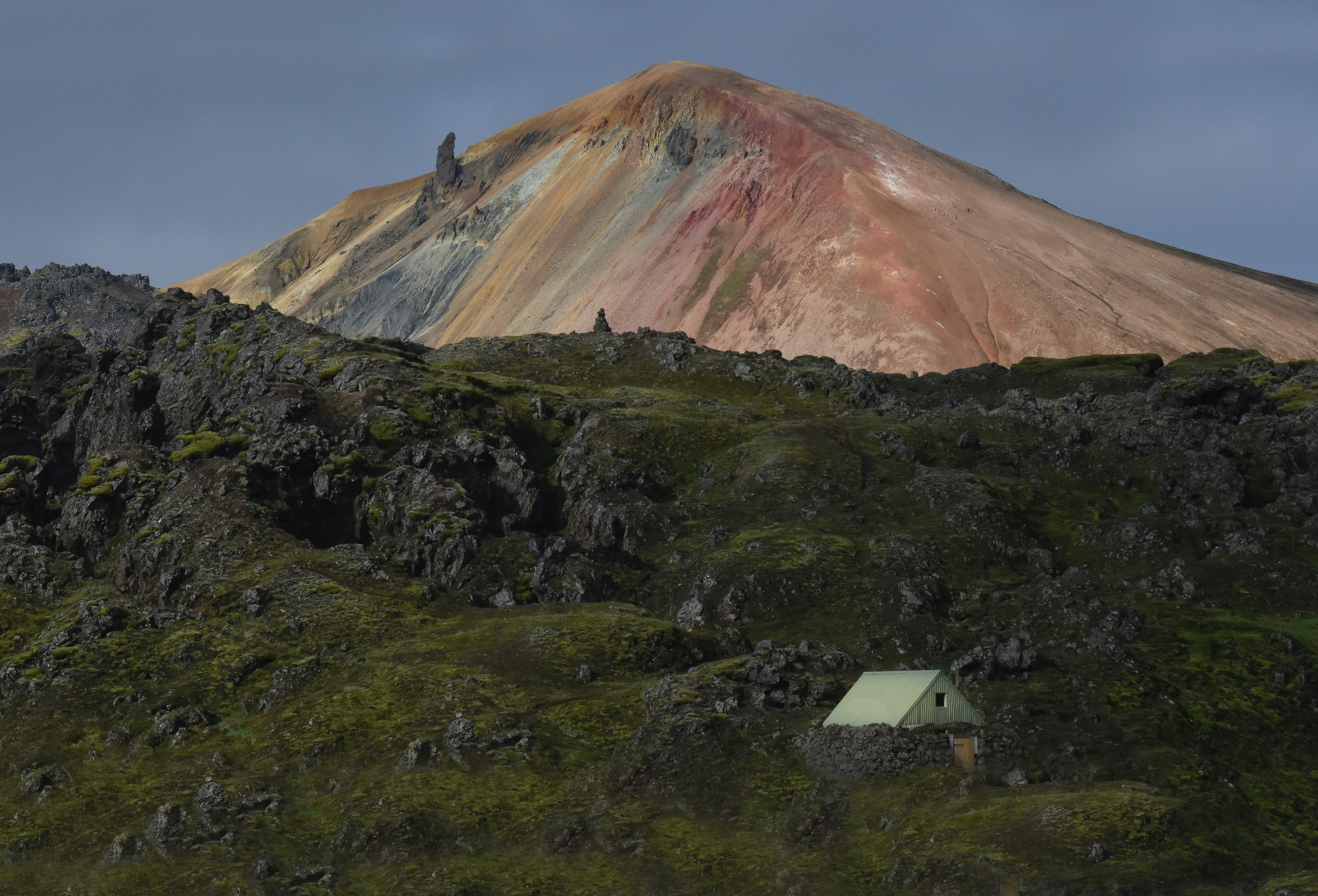 A small green cabin rests among rugged lava formations, with a colorful volcanic mountain looming in the background. The scene captures the stark beauty of Icelandic landscapes.