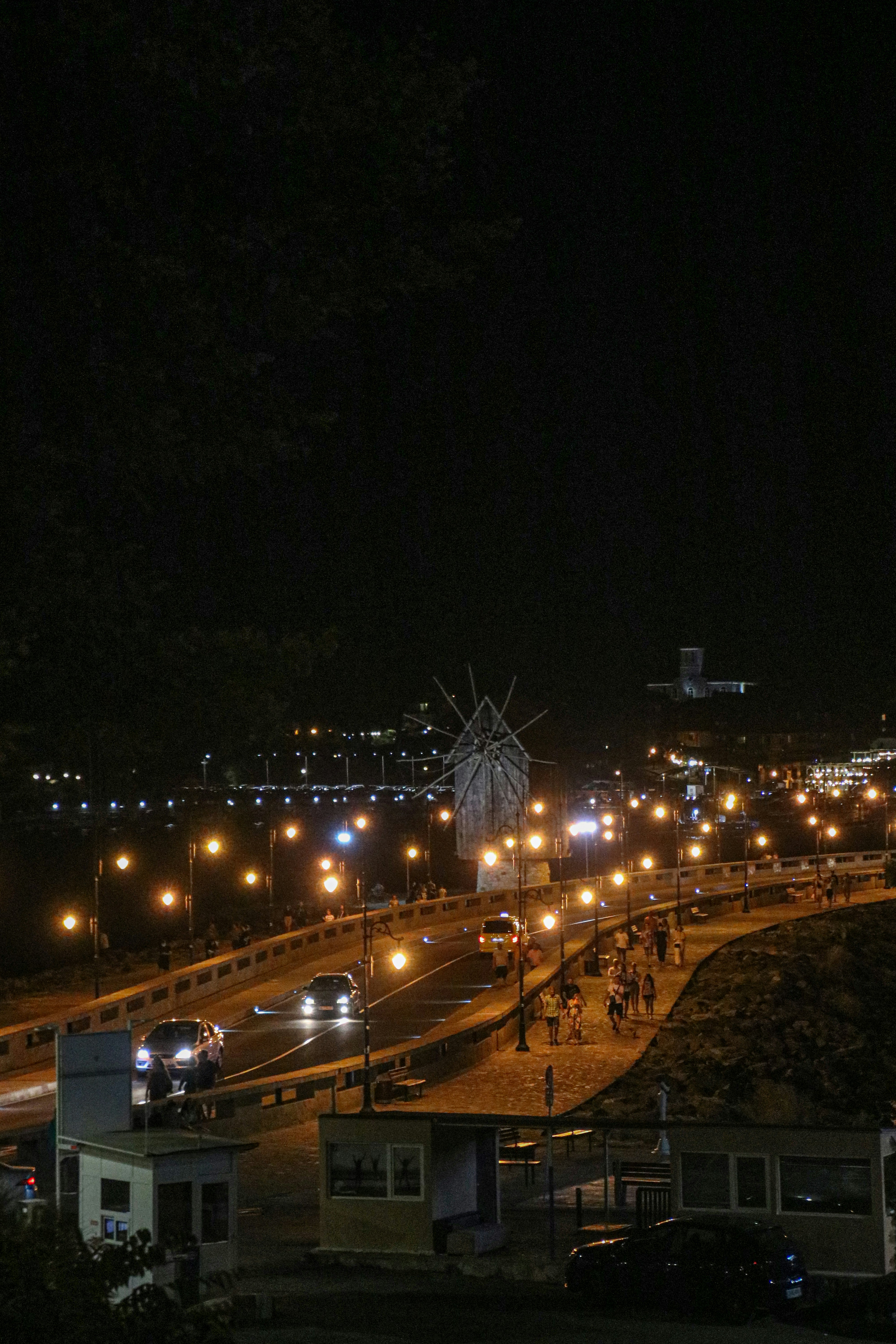 A vibrant riverside scene at night, showcasing illuminated pathways and a ferris wheel in the distance. The bustling activity of pedestrians and vehicles adds life to the tranquil waterside atmosphere.