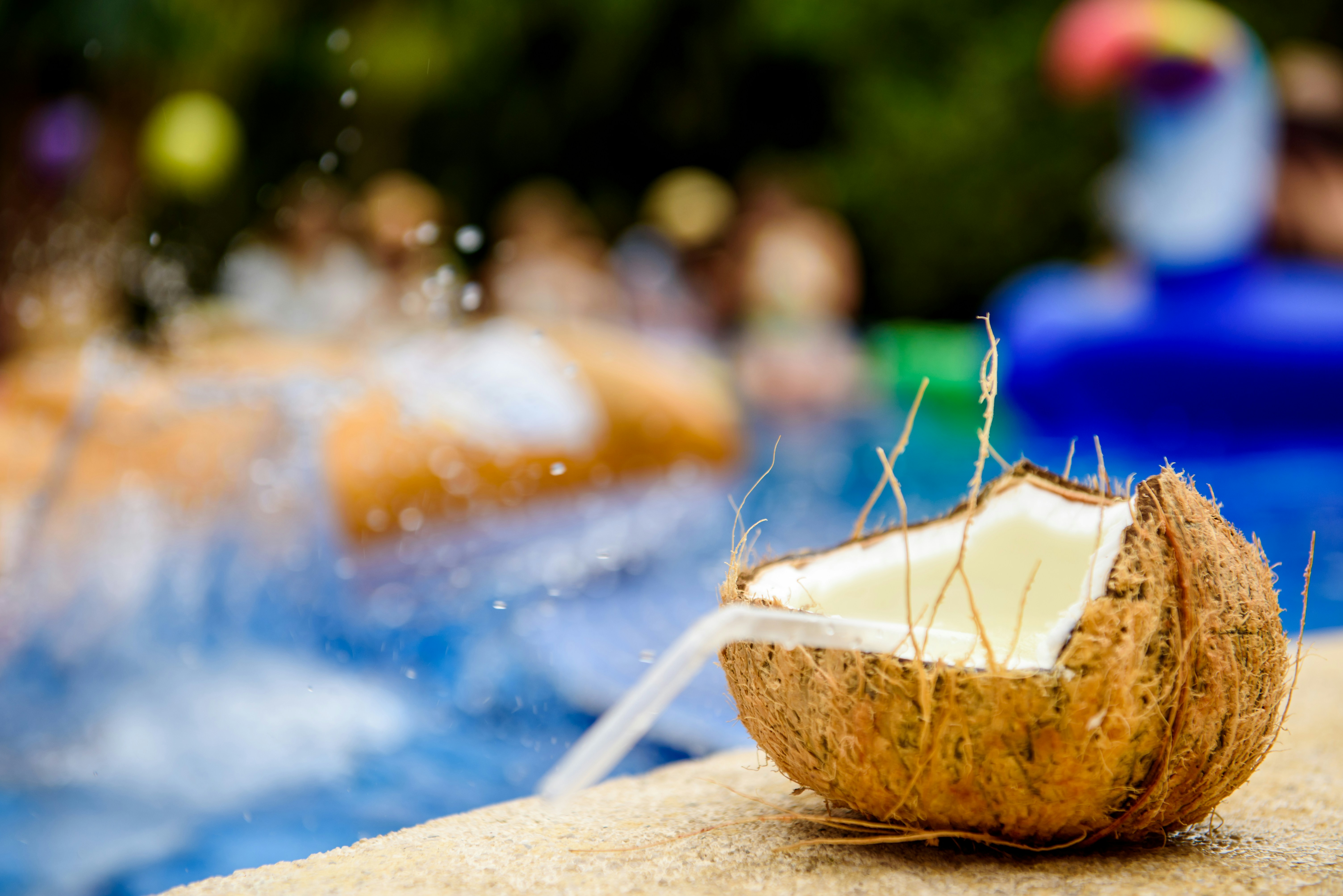 Freshly cracked coconut with water spilling out, set against a blurred pool backdrop with colorful floats. 