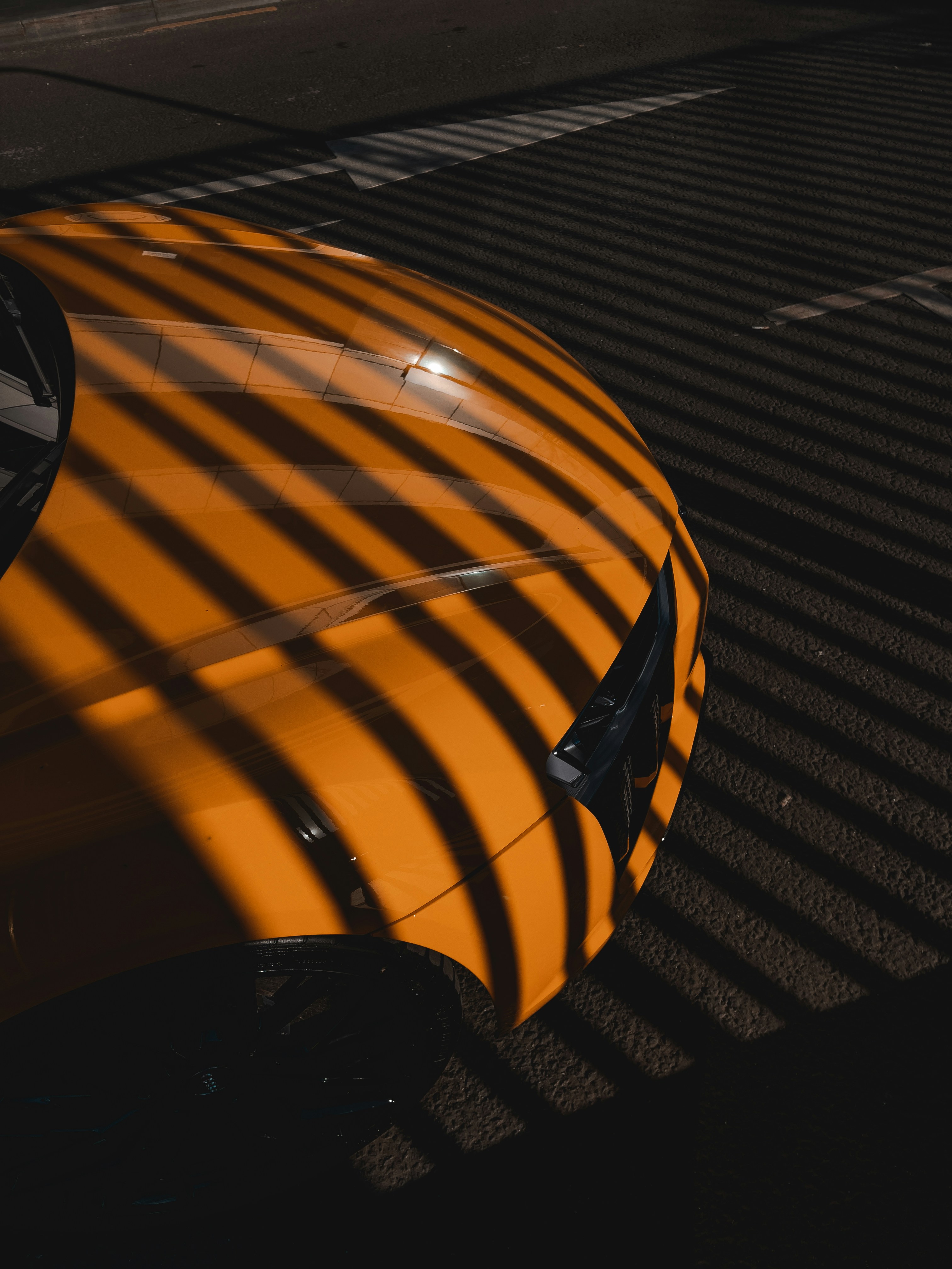Bright orange car partially obscured by dramatic shadows cast by striped patterns. The interplay of light and shadow creates a striking visual effect.