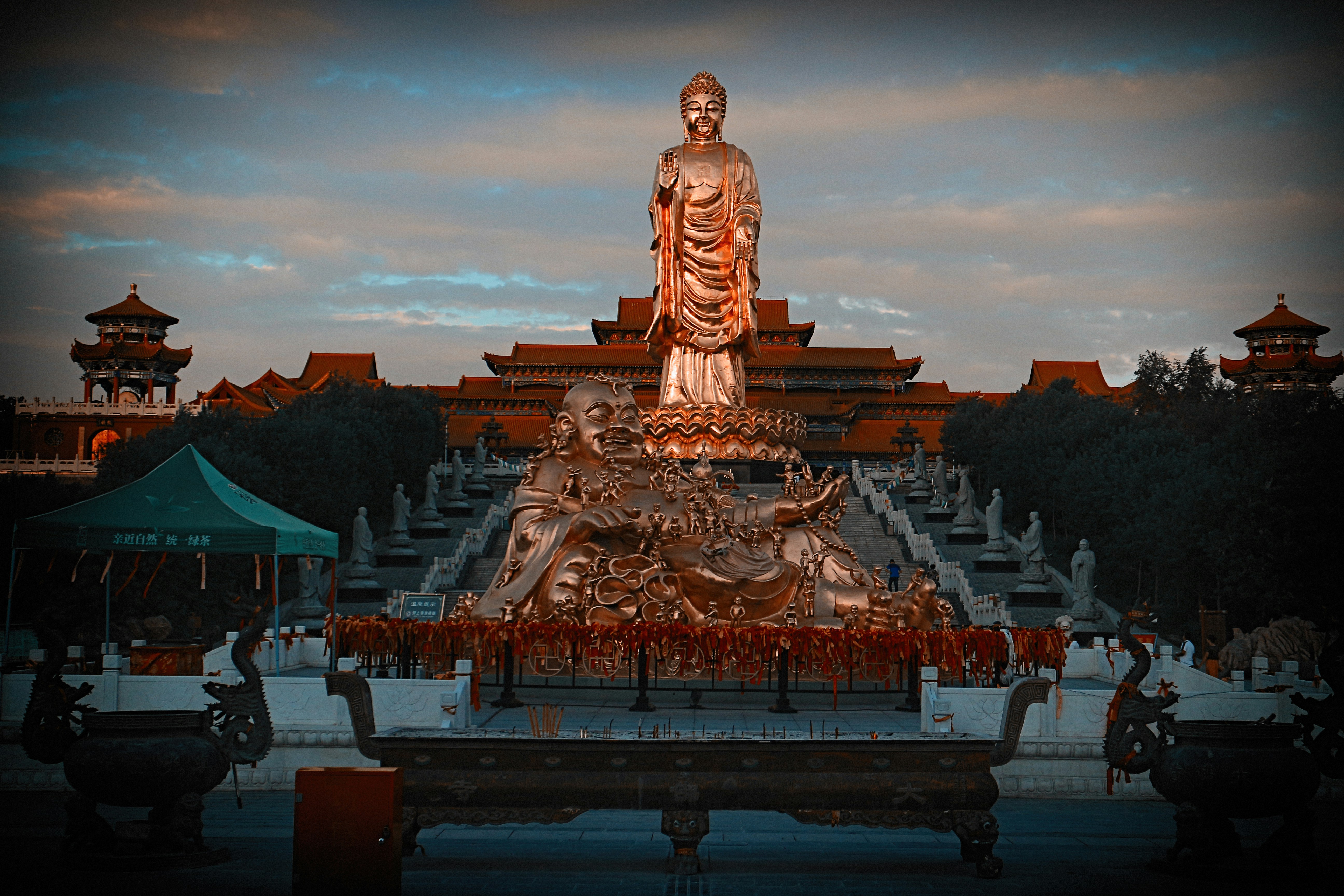 Statue of a robed figure on an ornate pedestal surrounded by traditional architecture under a twilight sky.