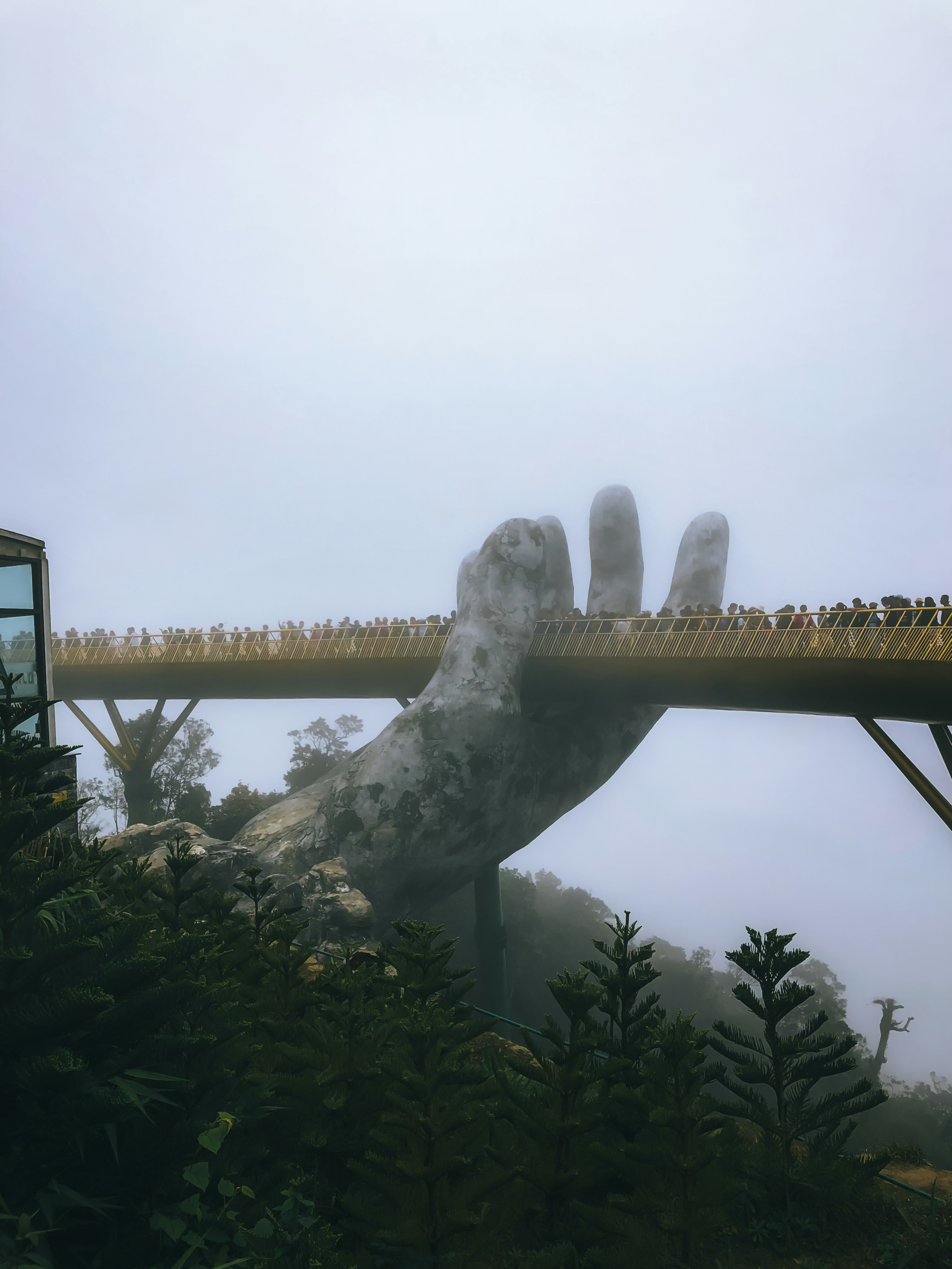 A colossal stone hand supports a bridge adorned with visitors, enveloped in misty surroundings. Lush greenery frames the scene, enhancing the ethereal atmosphere.