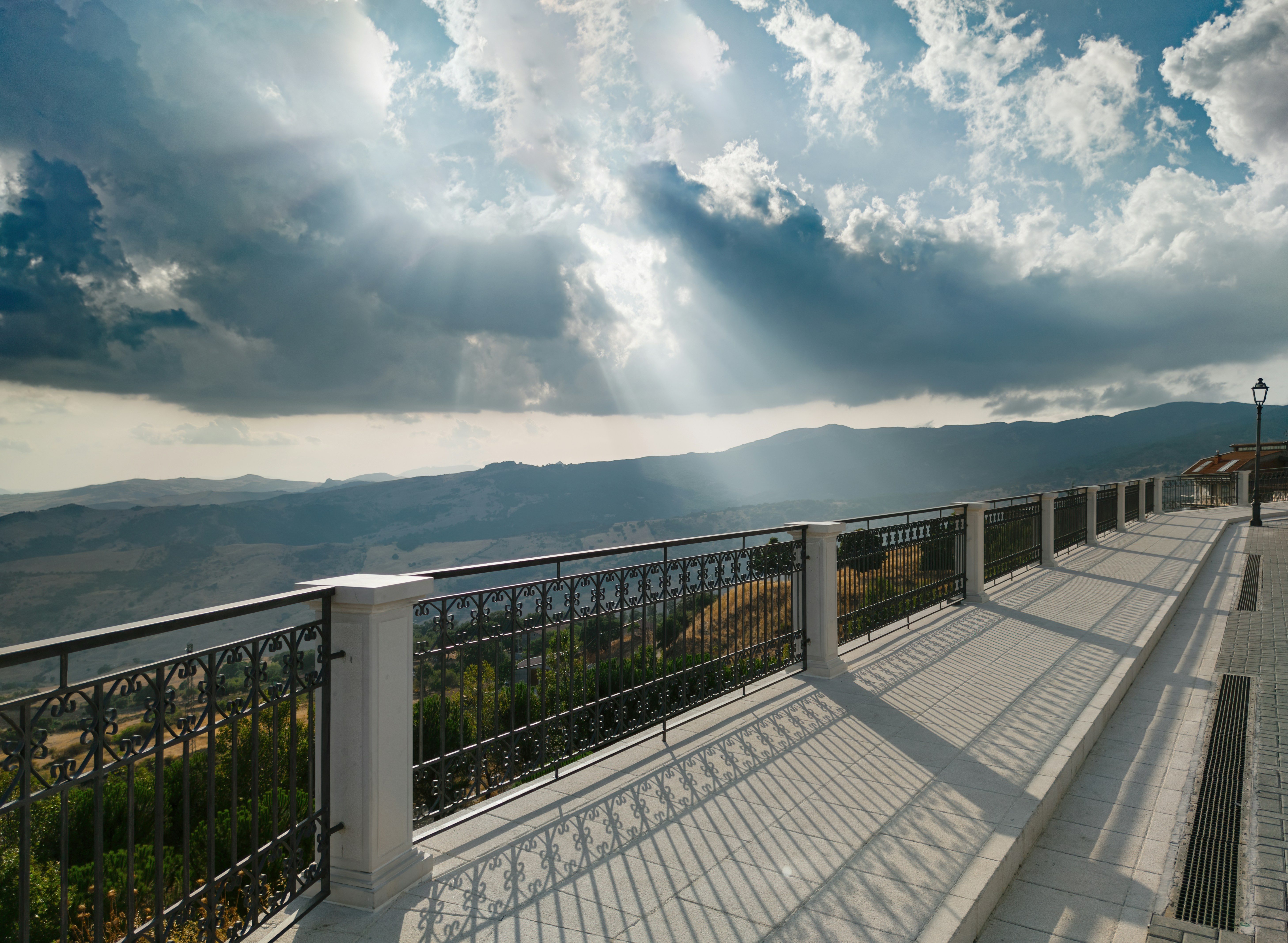 Photograph of a marble terrace with white columns and a metal railing, extending toward distant alpine ridges as sunbeams break through dramatic clouds.