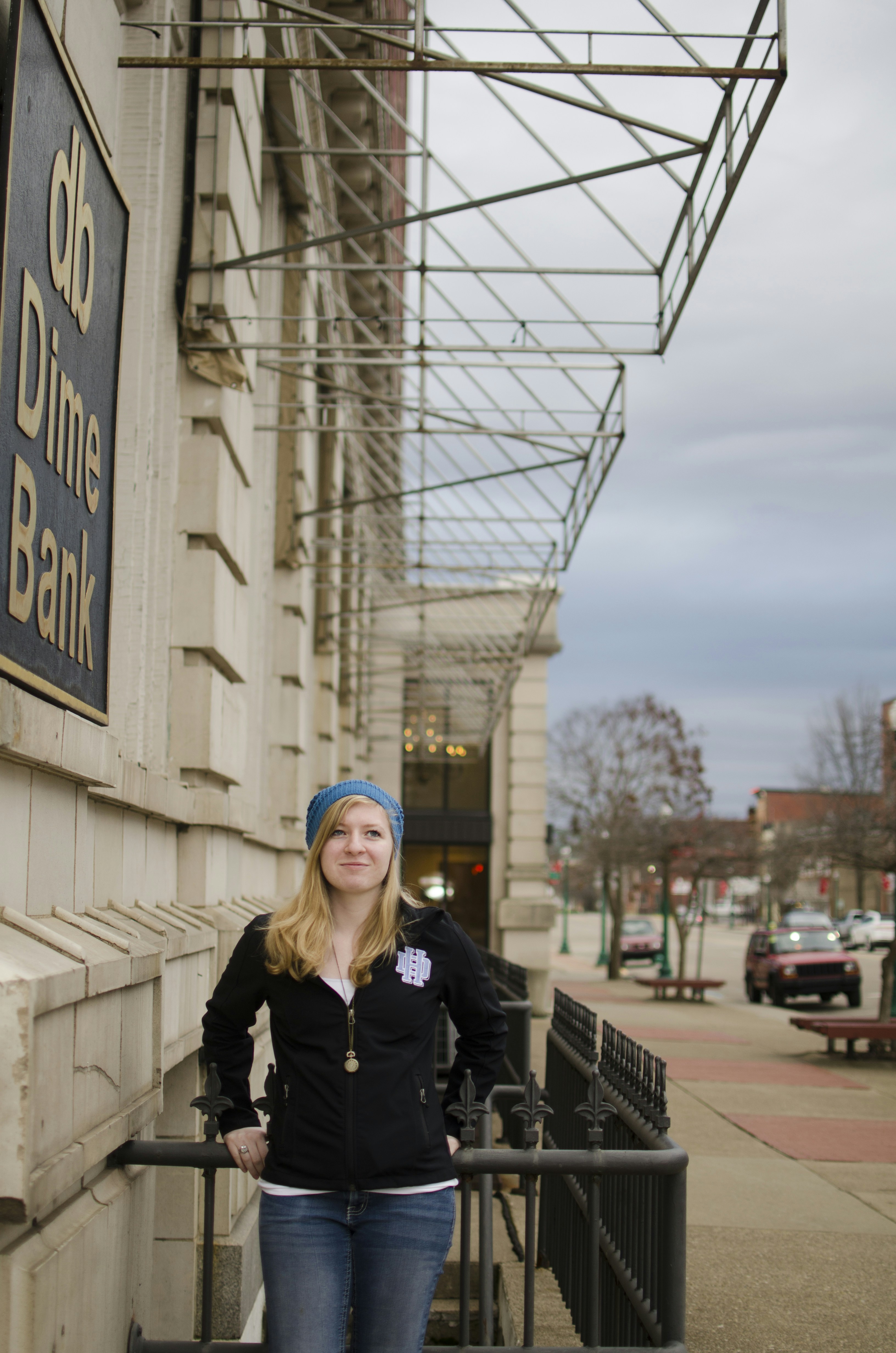 Street-front Portrait: A Blonde Woman in a Blue Beanie at Dime Bank Lounges by the Sidewalk
