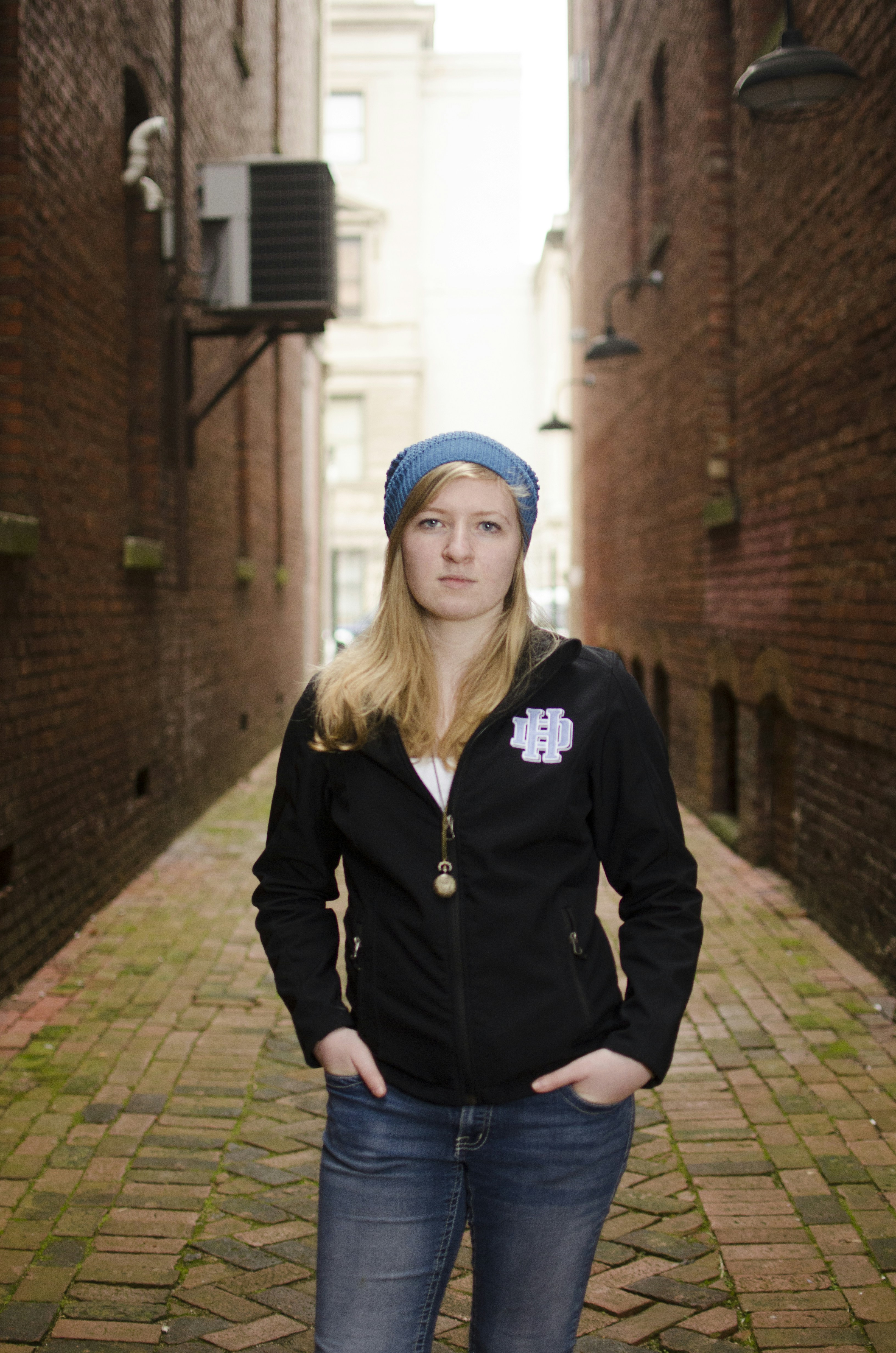 urban female senior photo poses blonde hair blue beanie outdoor city small town | woman in black jacket and blue knit cap standing on brick pathway during daytime