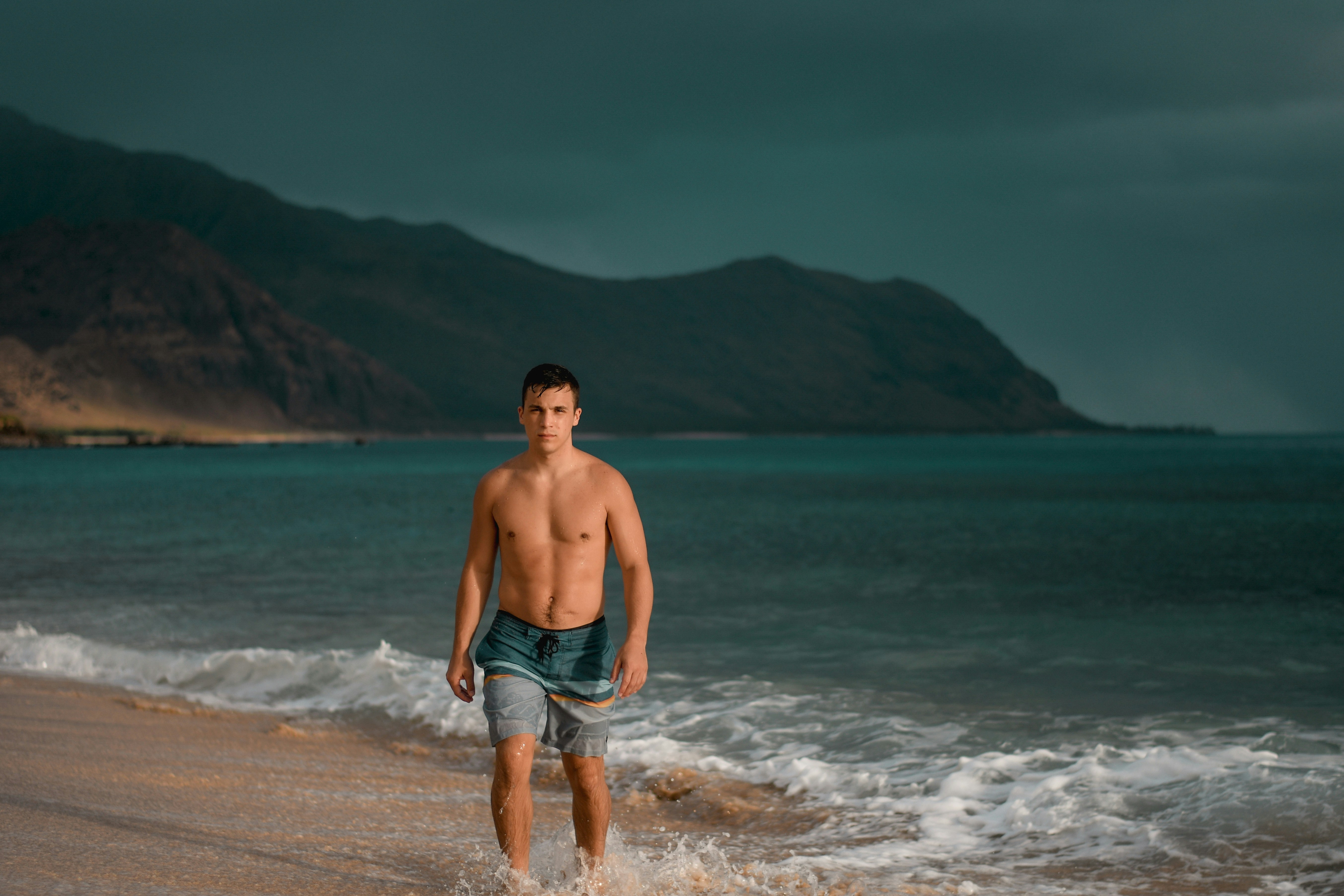A young man strides through gentle waves on a beach, framed by dramatic mountains under a moody sky.