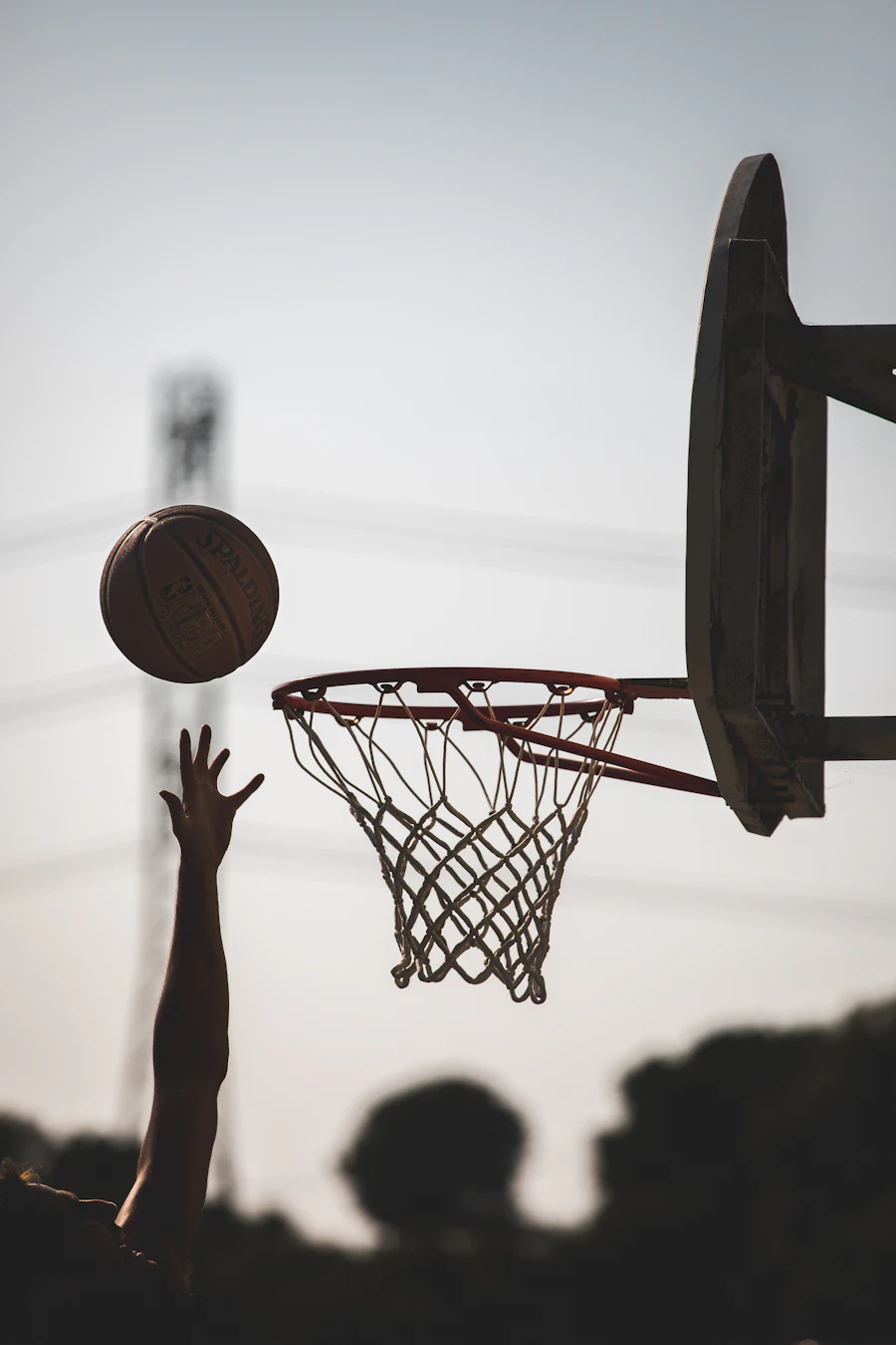 Basketball hovering above a hoop as a player's outstretched hand reaches toward the rim at dusk.