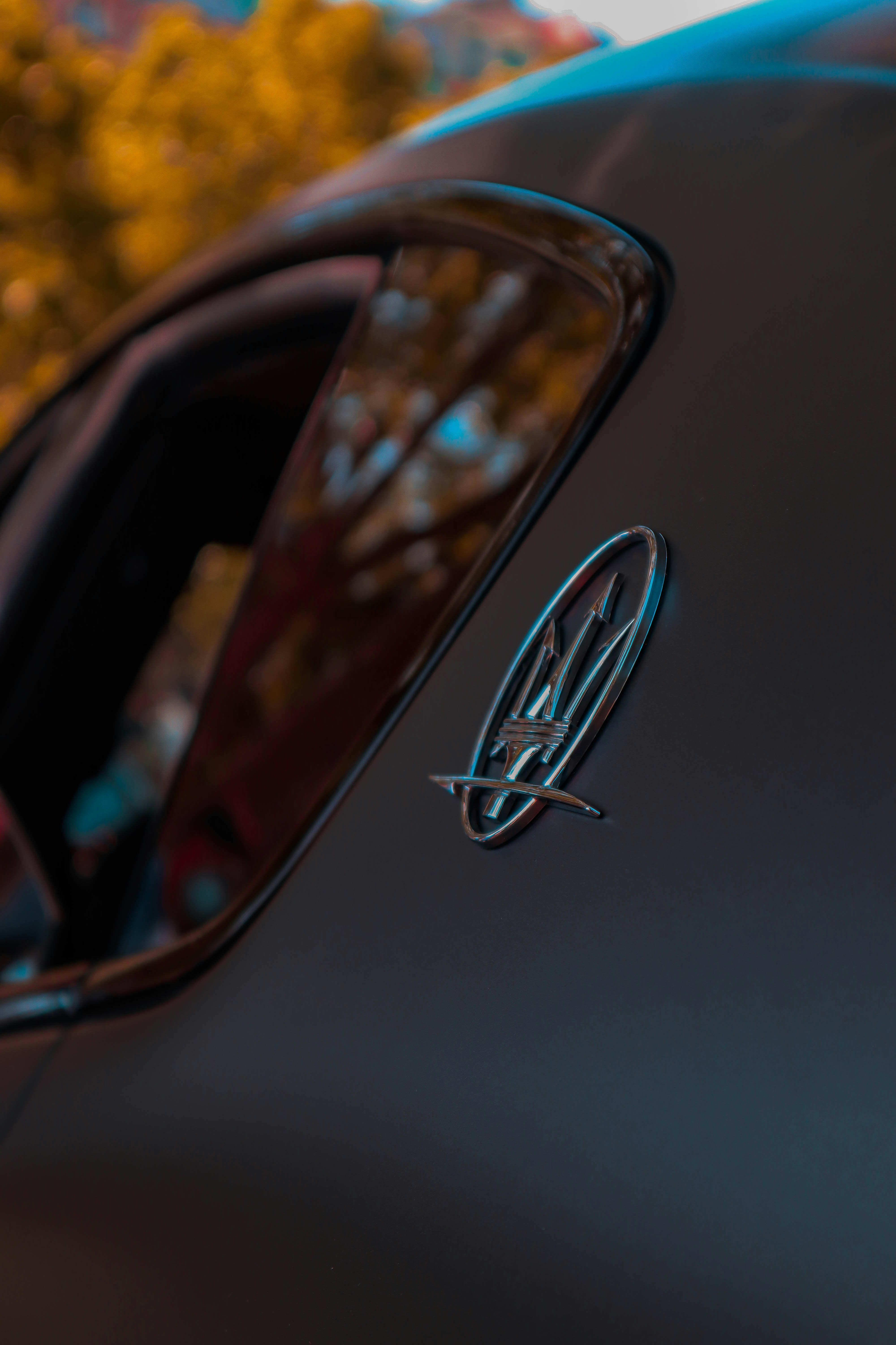 Close-up of a Maserati emblem on a sleek vehicle, highlighting its luxurious design against a blurred backdrop of autumn foliage.