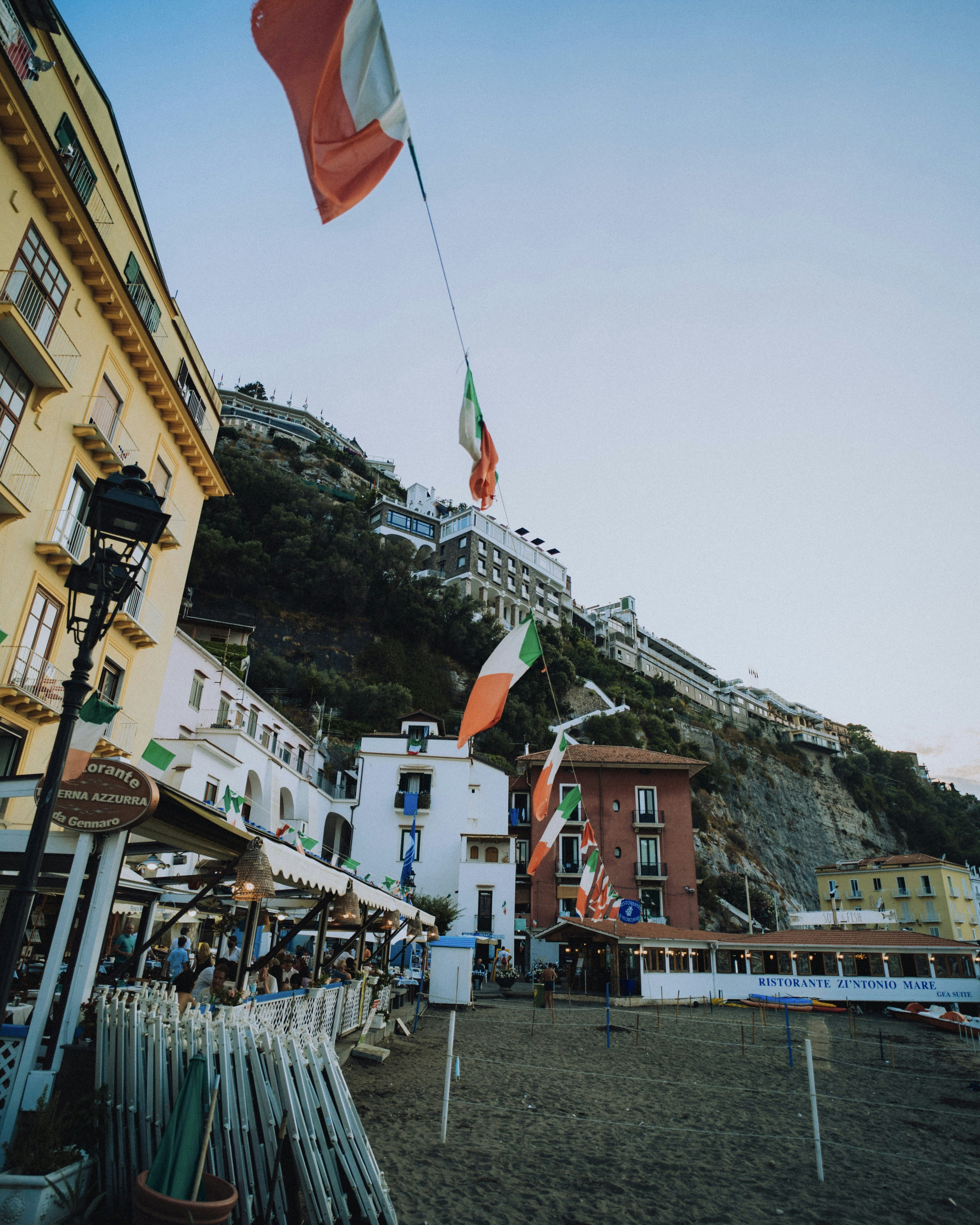 Colorful flags flutter above a bustling beachside scene with charming buildings and a hillside backdrop. The lively atmosphere invites visitors to explore the coastal beauty.