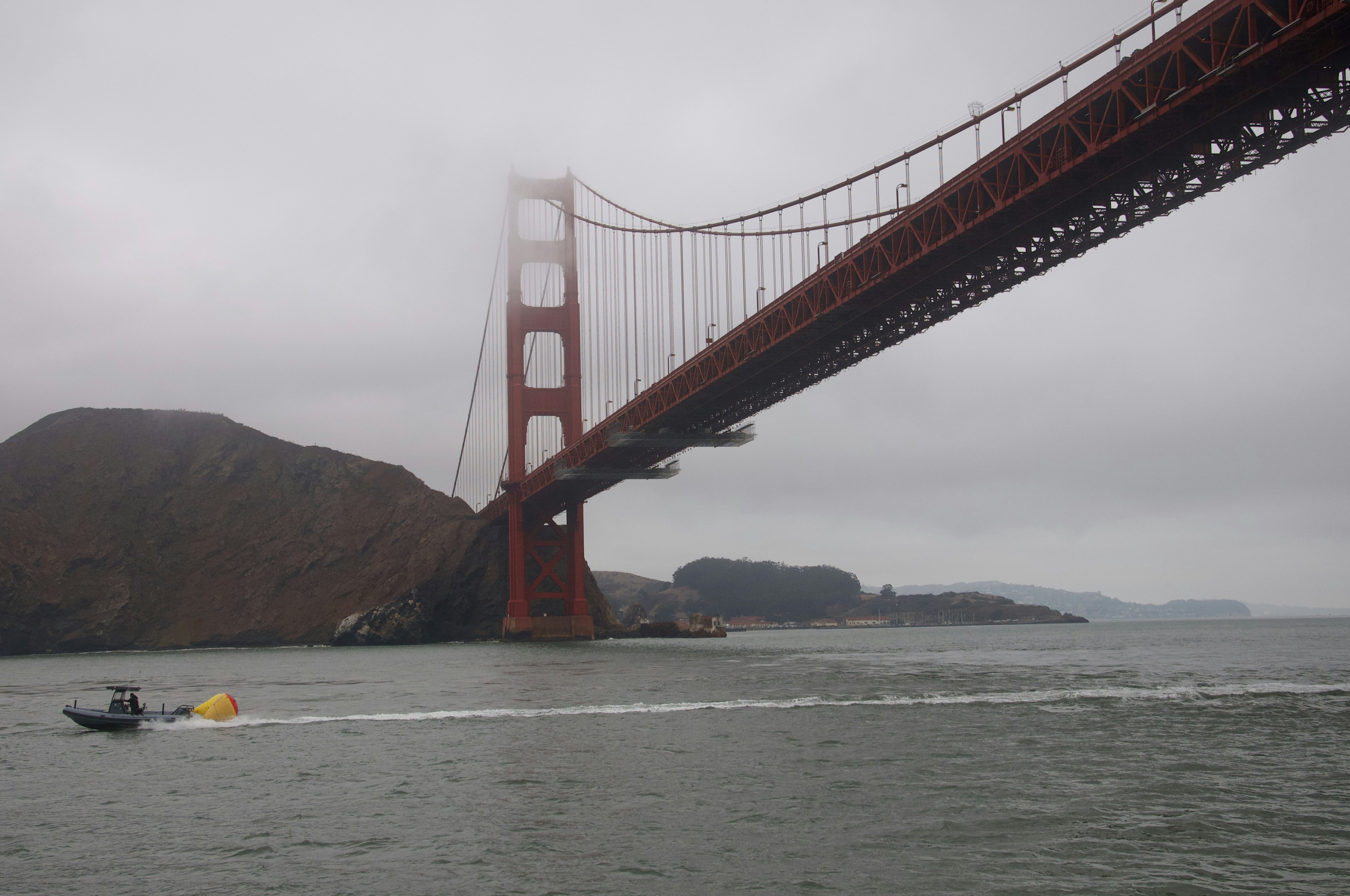 A boat glides through the waters beneath the Golden Gate Bridge, shrouded in fog, with rocky landforms in the background.