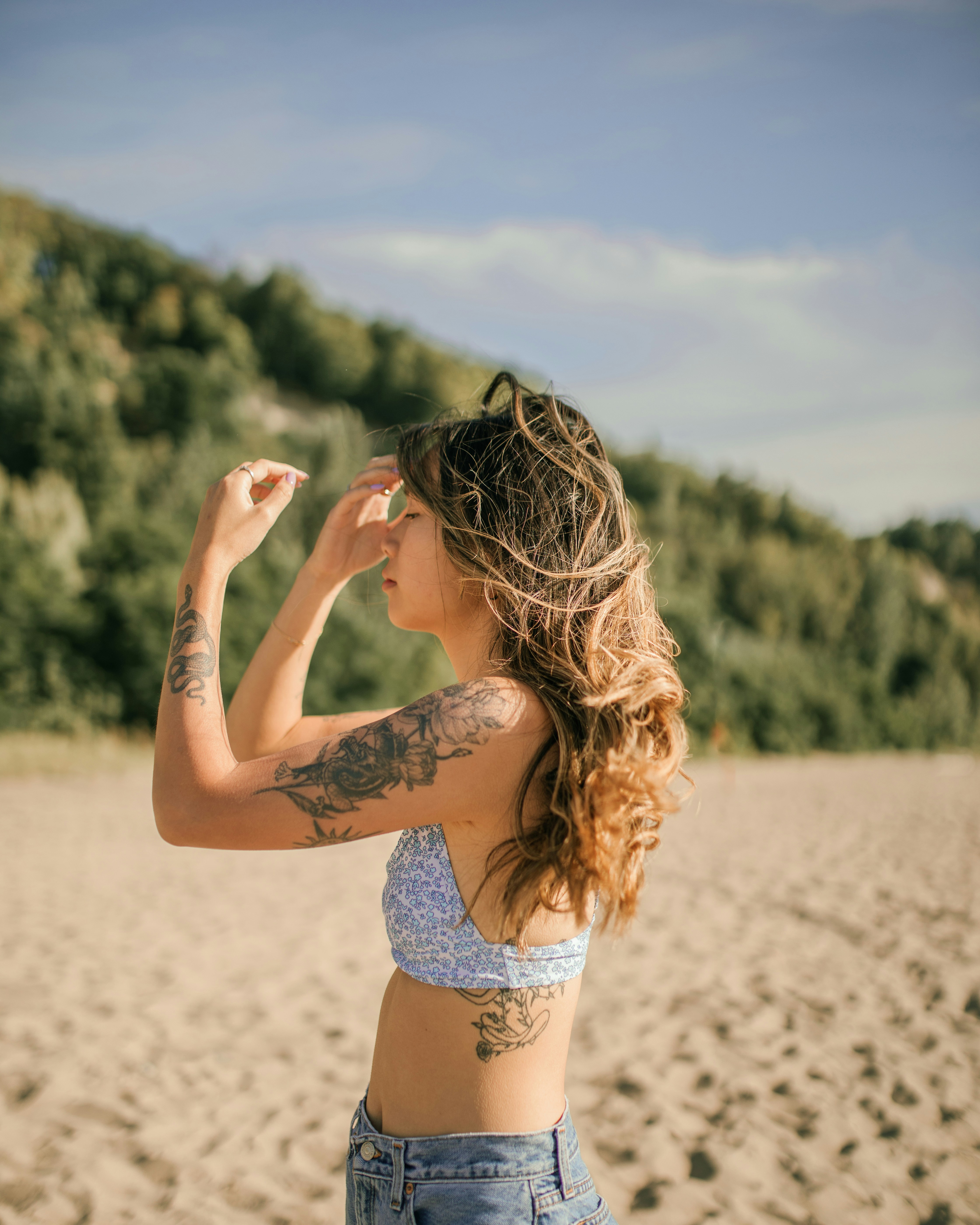 A woman with flowing hair and tattoos stands on a sandy beach, shielding her eyes from the sun while surrounded by lush greenery.