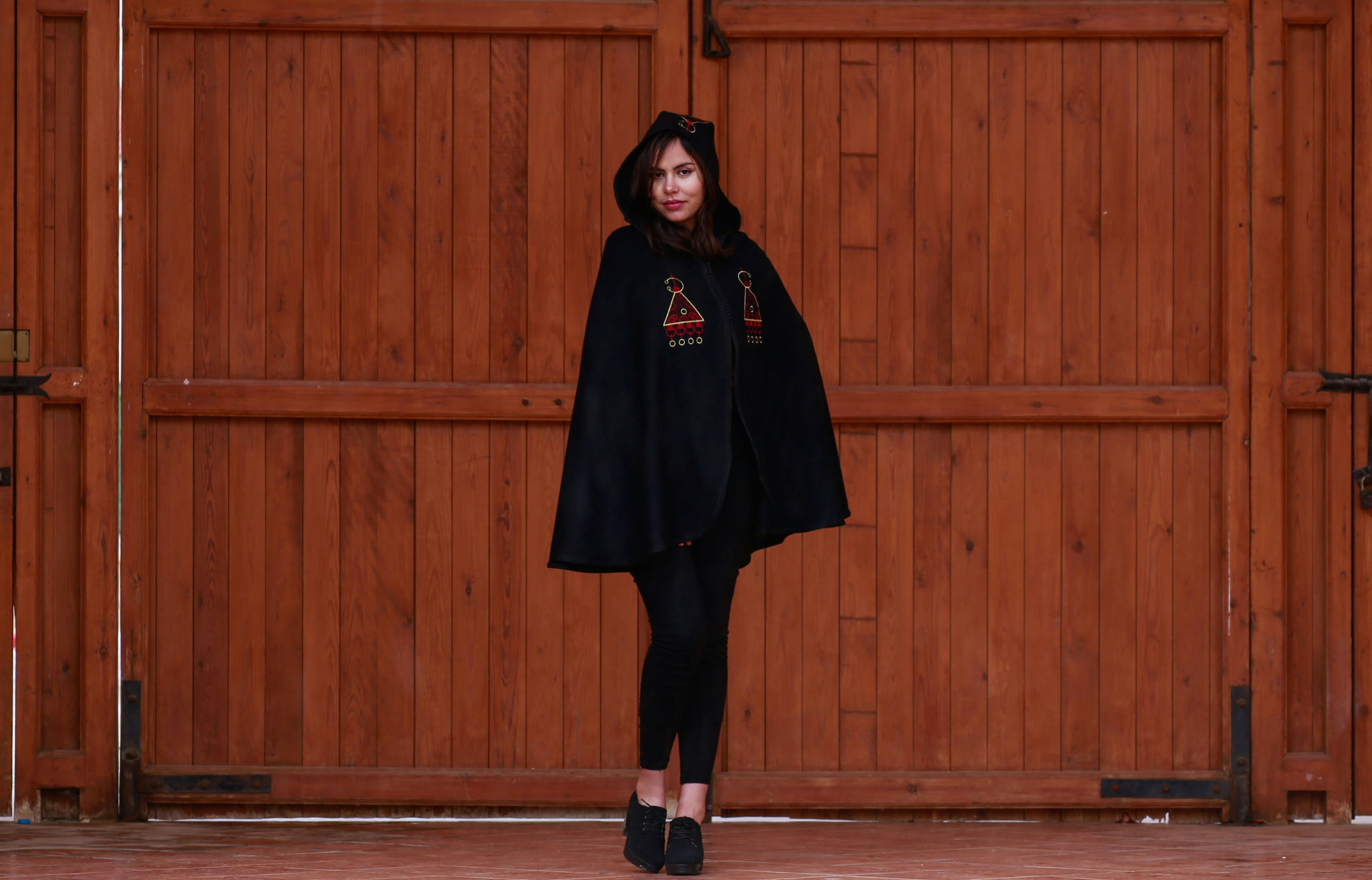 A figure in a dark cape stands confidently against a rustic wooden backdrop, exuding an air of mystery and elegance.